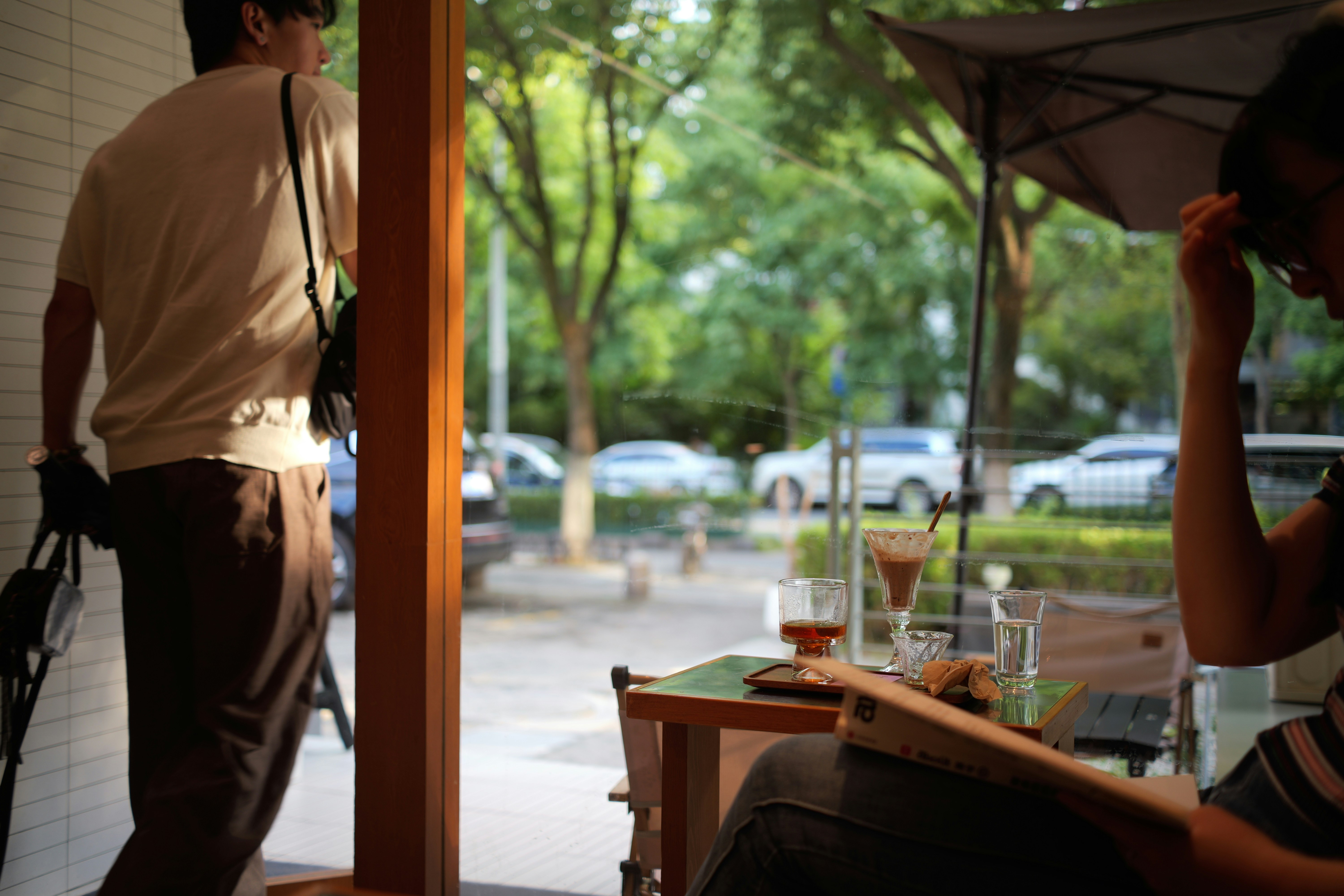Sunlit café scene with an open doorway to a tree-lined street; foreground shows a table with drinks and a person reading, while another figure sits off to the side. This is a candid interior shot from a seated perspective.