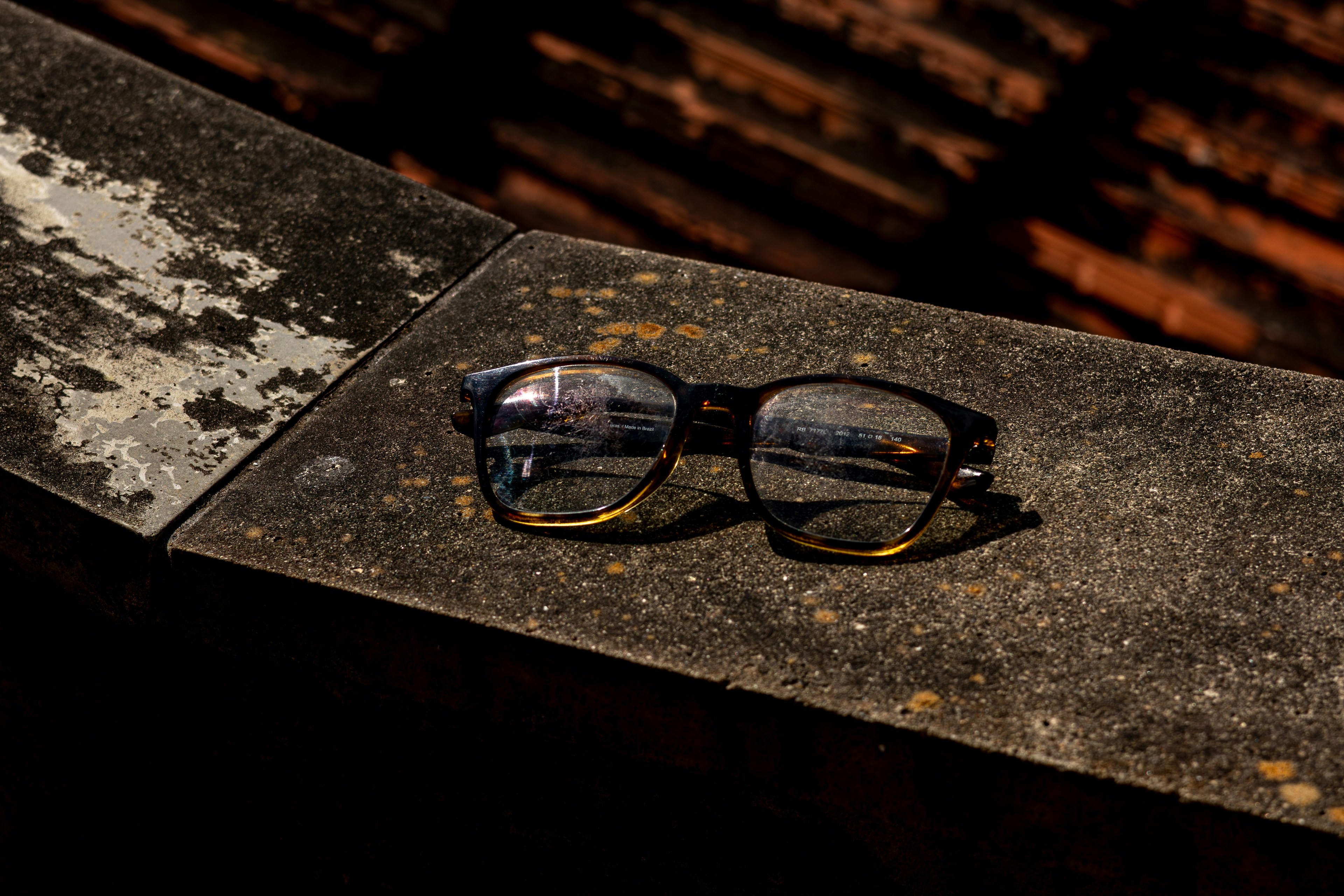 A pair of glasses rests on a rough concrete ledge, catching light and casting subtle reflections; the scene emphasizes texture and a solitary object against a dark background. This is a photograph, not a drawing or painting.