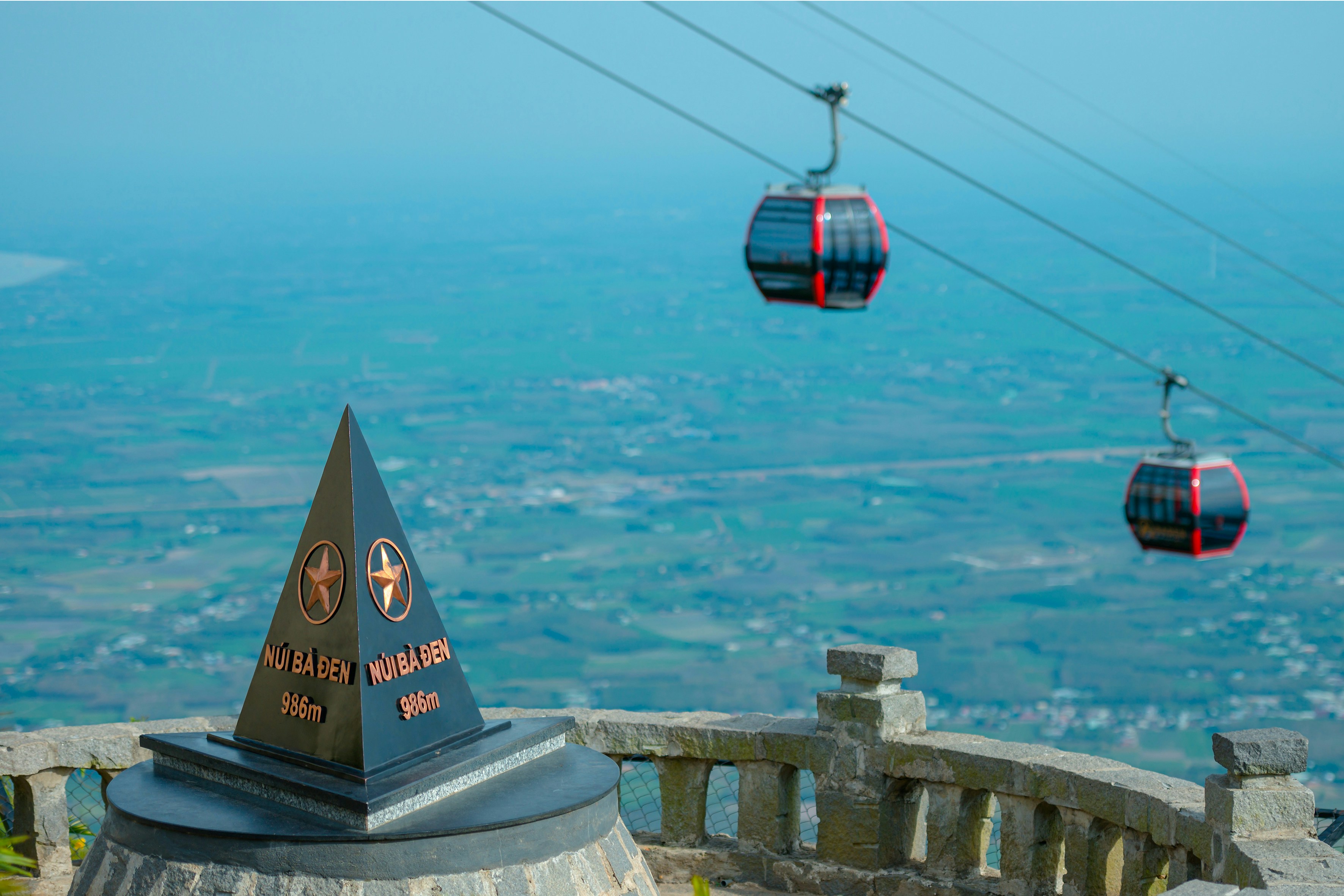 A summit monument and stone railing sit in the foreground with suspended cable cars gliding above a expansive valley below.
