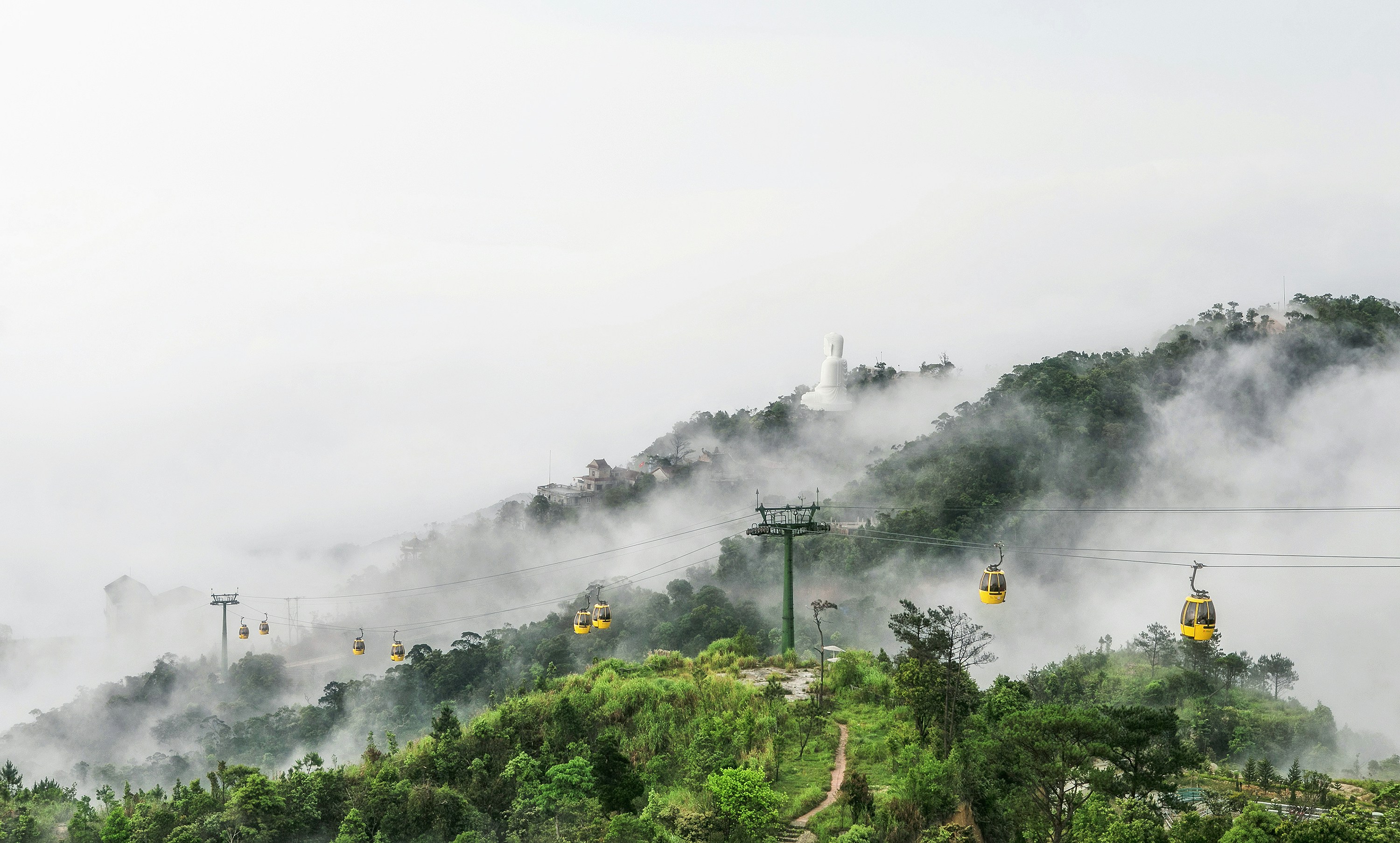 Cable cars glide through mist over a green hillside, with a distant white structure atop the ridge.
Note: This is a landscape photograph capturing fog, vegetation, and a funicular-like cable system.