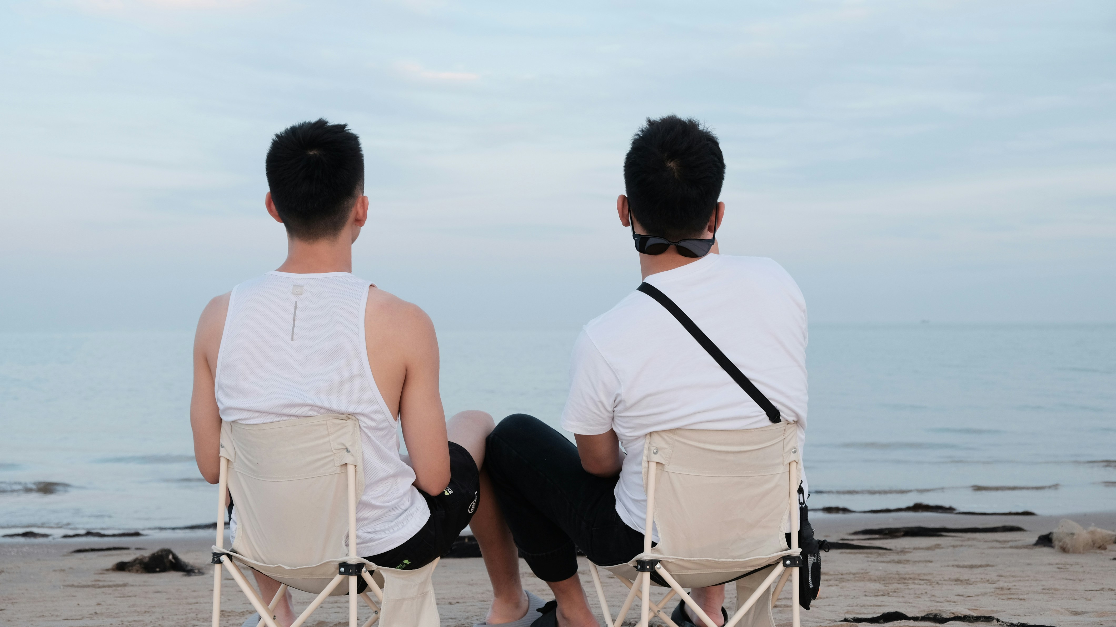 Two men in white shirts sitting on folding chairs on a sandy beach