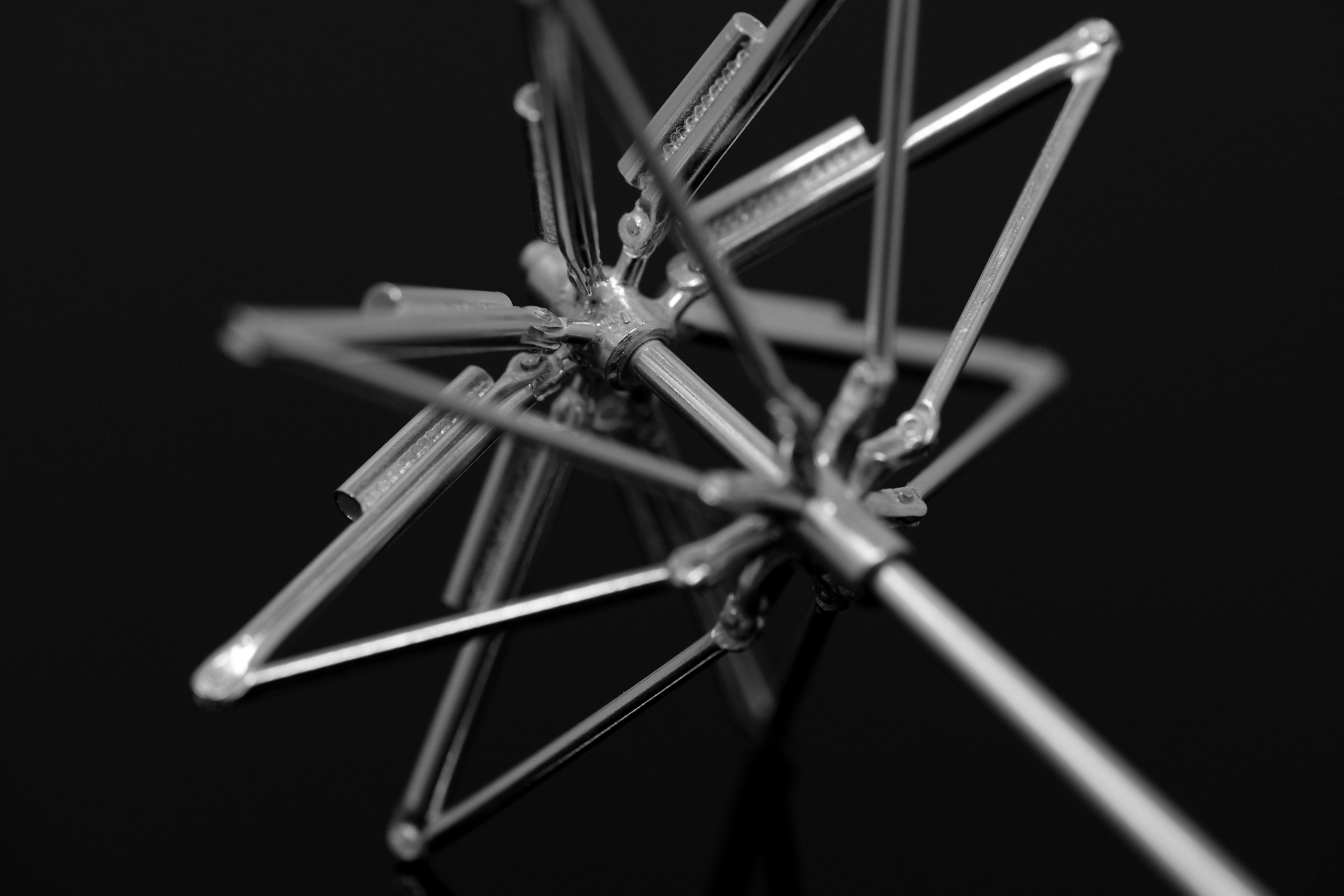 Close-up of a metallic geometric structure set against a dark background, capturing intricate intersecting rods and joints with high-contrast black-and-white tones. Abstract sculpture, no identifiable subject.