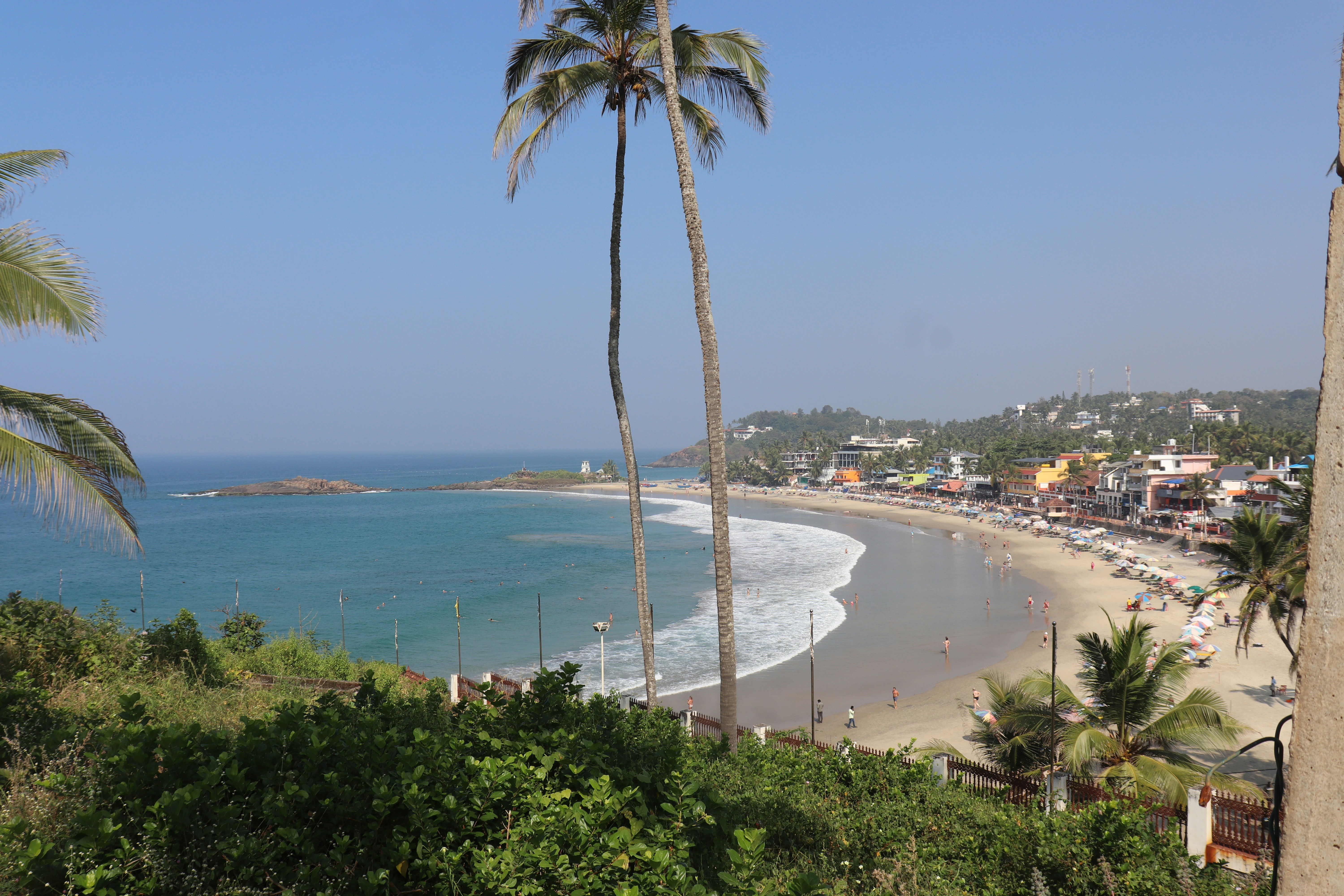 Sunlit coastal village curves along a sandy beach with palm trees in the foreground and a calm sea beyond. A panoramic view capturing shoreline activity and blue sky.