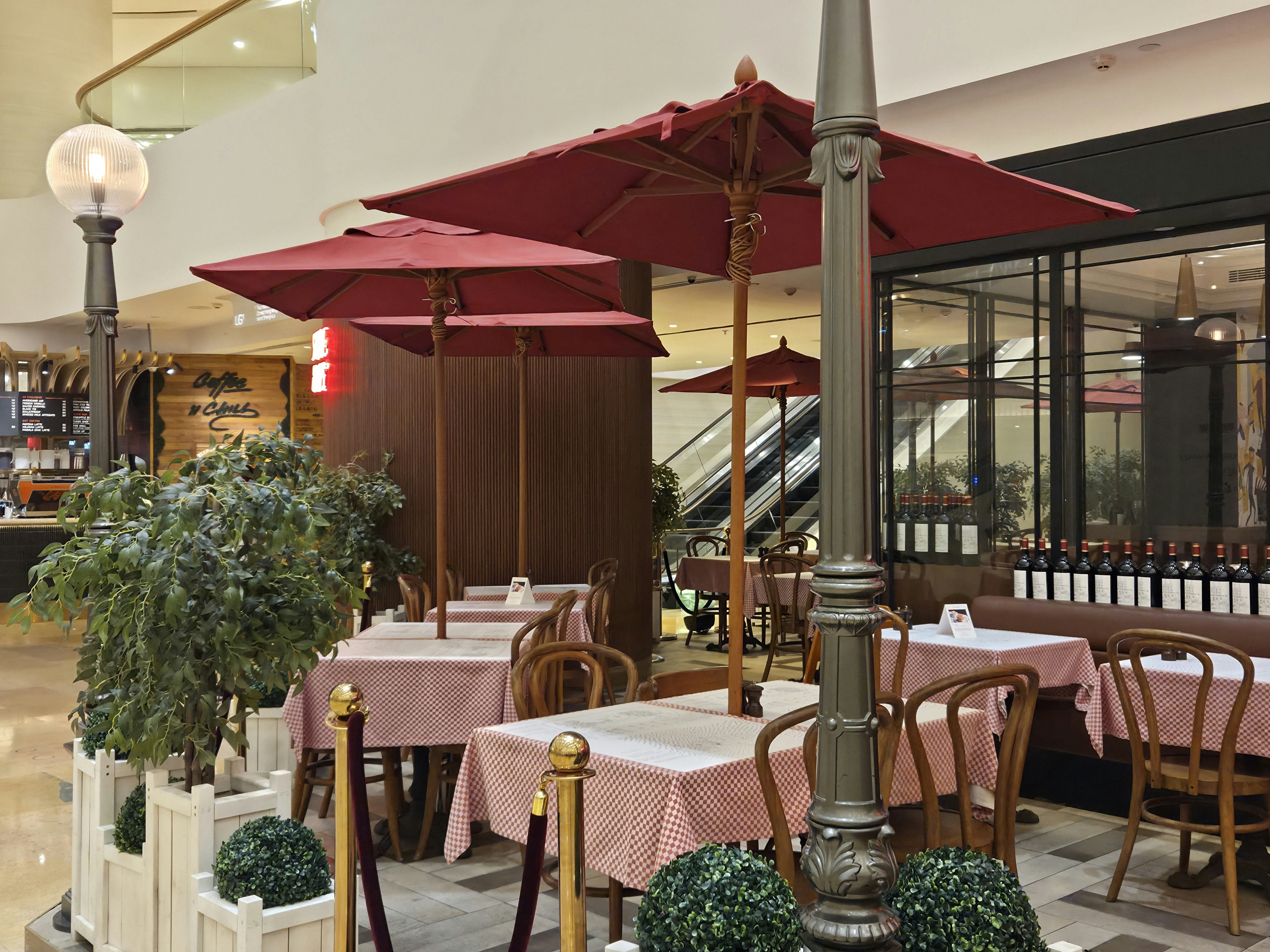 Warm cafe seating with checkered tablecloths under burgundy umbrellas in a modern shopping center courtyard.