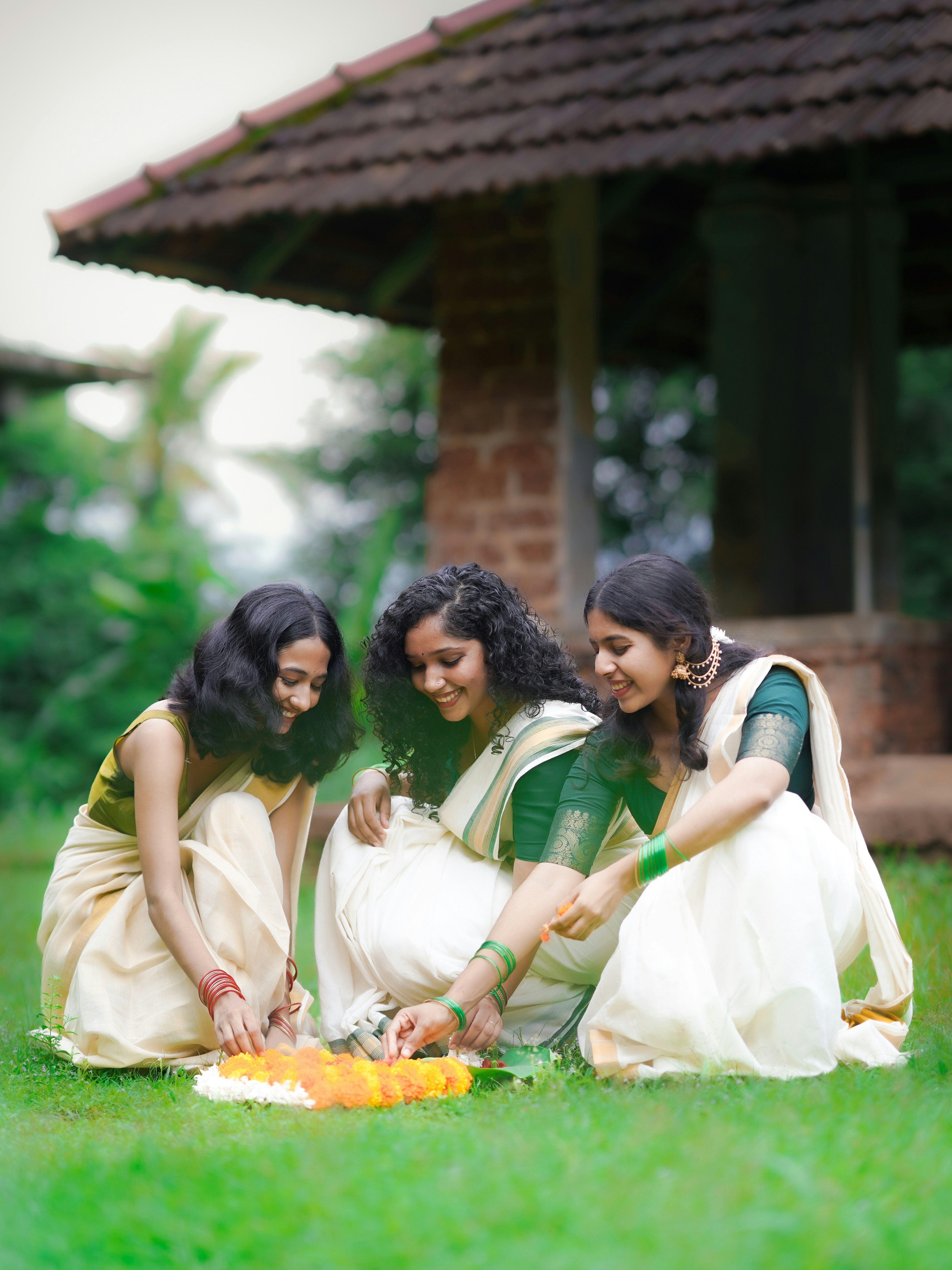Three Friends in Cream Sarees Gather Flowers on Lush Green Ground