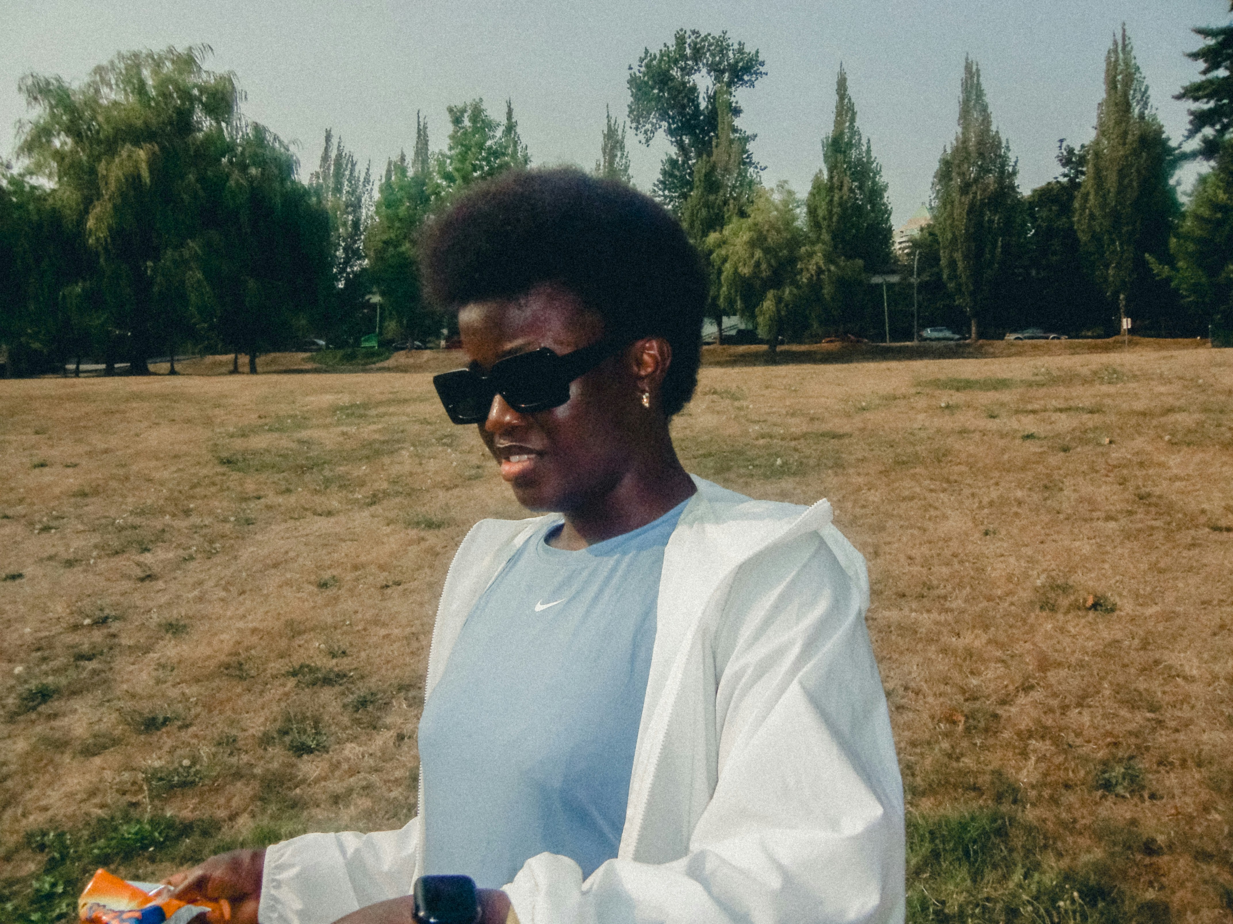 A woman in sunglasses and a light jacket in a dry grassy park