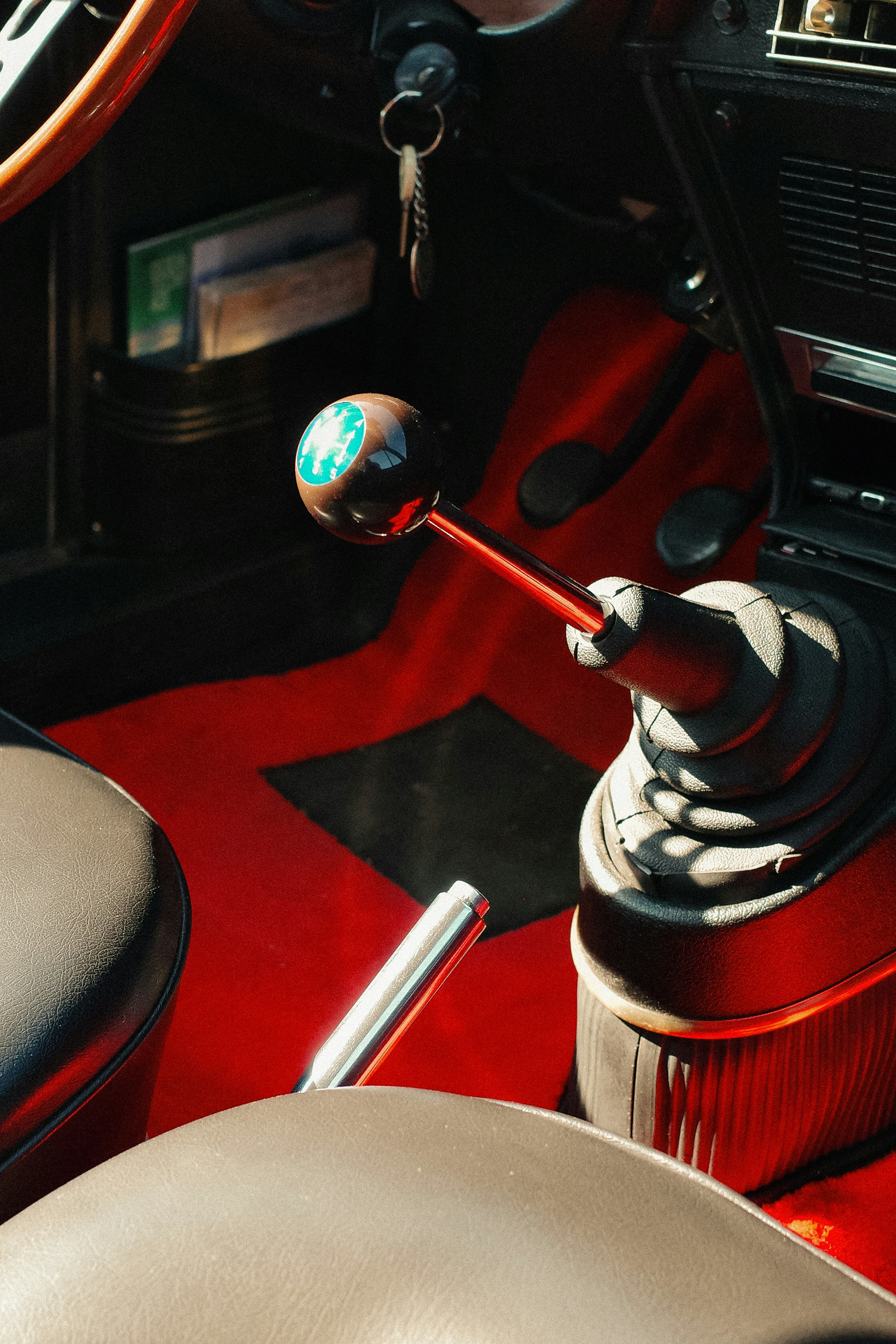 Close-up of a vintage manual gear shift in a car interior with red carpeting and a surrounding dashboard; the scene emphasizes the gear lever and surrounding controls.