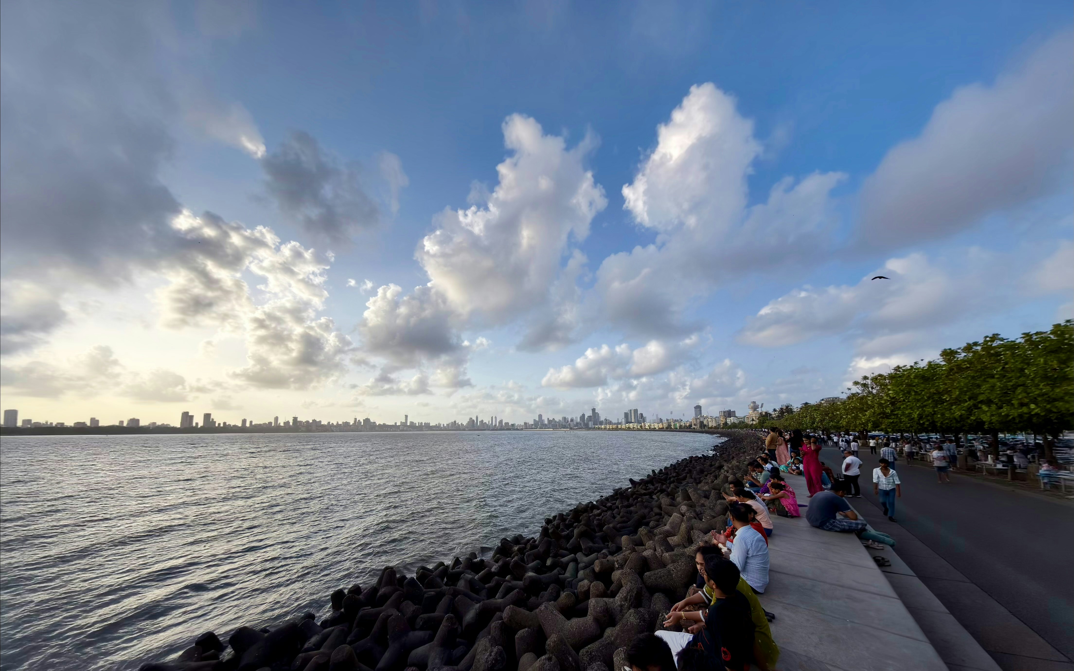 Crowd seated along a seawall by a wide river, city skyline on the horizon with a rocky jetty and a tree-lined promenade. A calm, daytime urban riverside scene showing people and open sky.