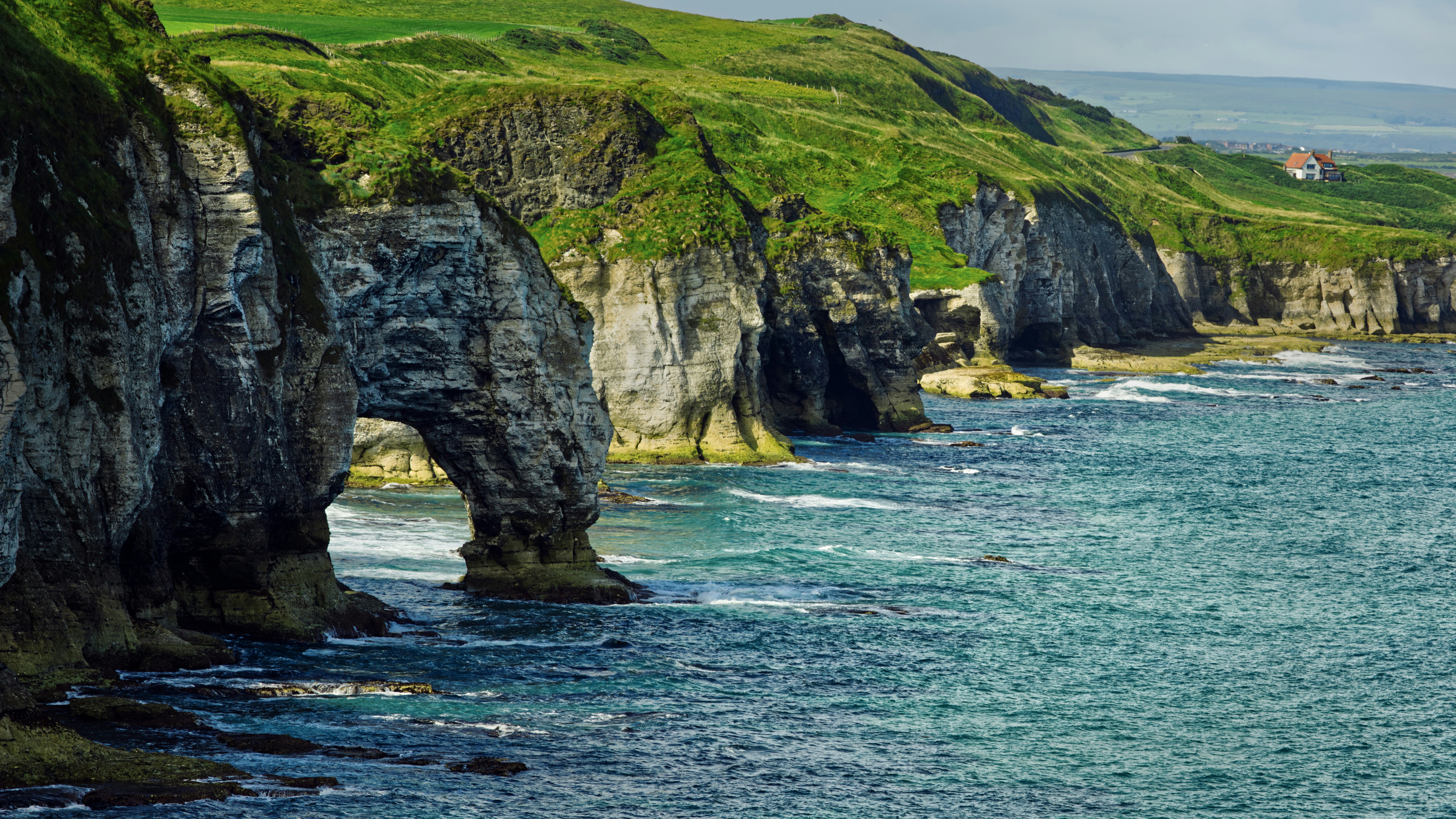 Coastal cliffs with a natural sea arch rising above turquoise water, grassy top edge, and a distant house on the horizon. A wide landscape scene emphasizing rugged geology and sea spray.