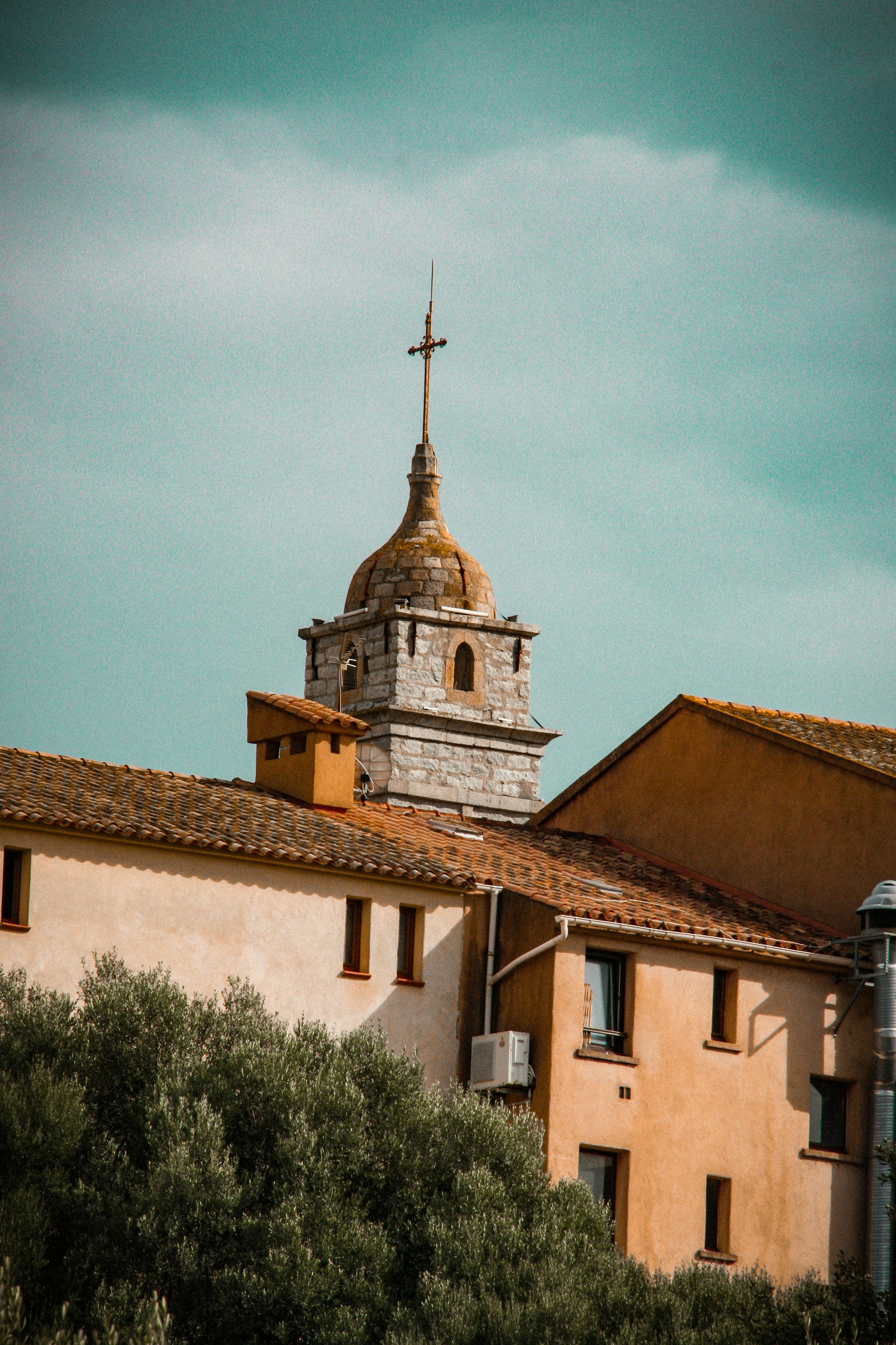 A stone church bell tower with a cross above terracotta roofed buildings