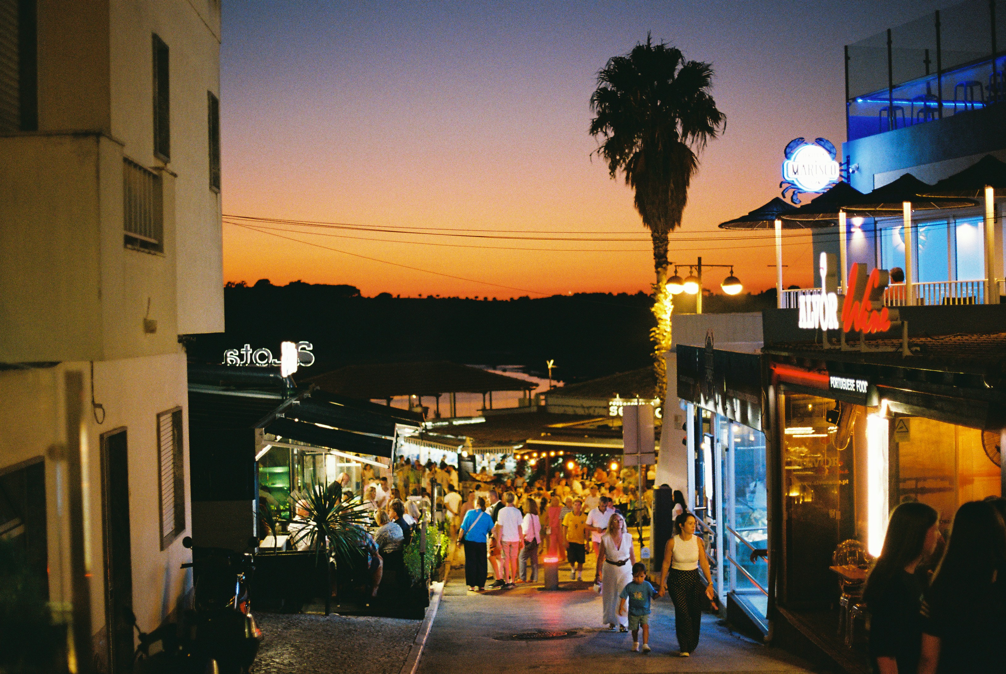 Bustling market street at dusk with a glowing palm silhouette and colorful storefronts.