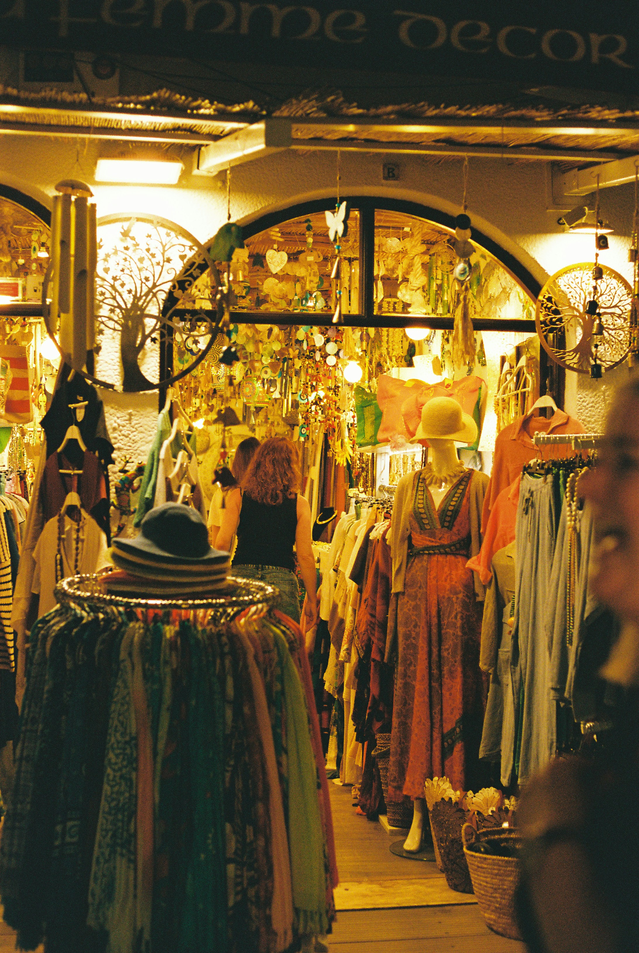 A warmly lit boutique filled with racks of clothing and decorative items; a central hat display foregrounds the scene with a long aisle of dresses and shoppers in the background.