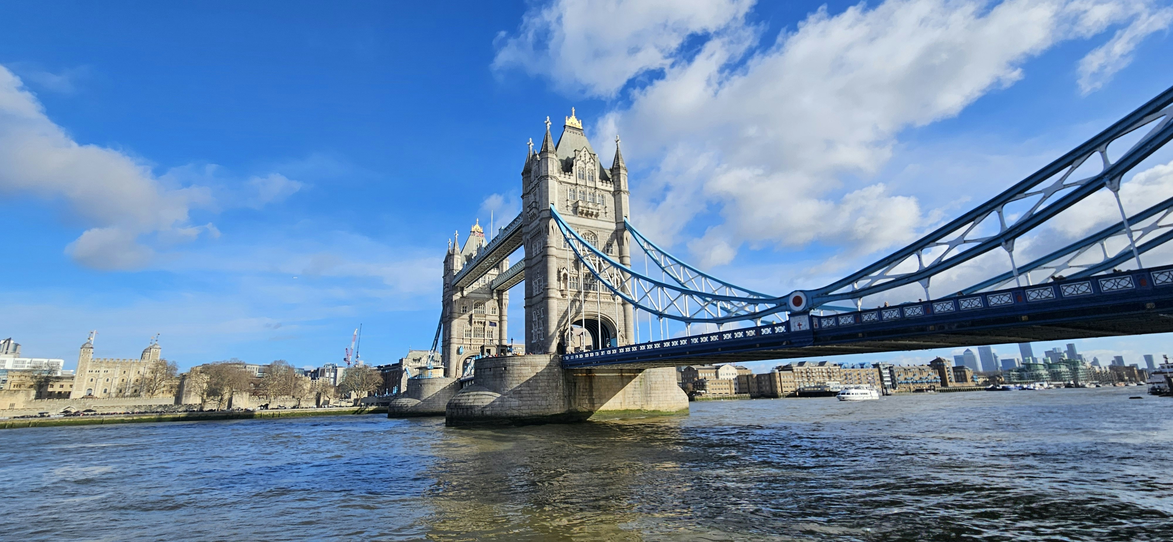 Tower Bridge rises over the River Thames with a bustling riverside and a blue sky above.