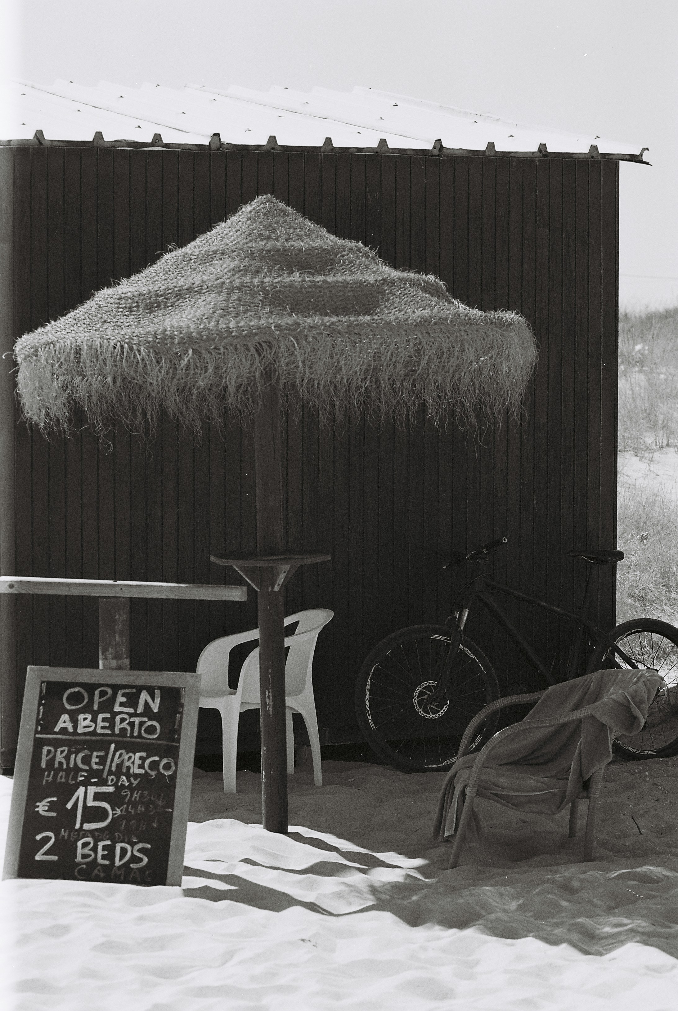 Stripey hay parasols hang above a sand-covered scene in front of a dark shack, with a chalkboard sign and a bicycle nearby.