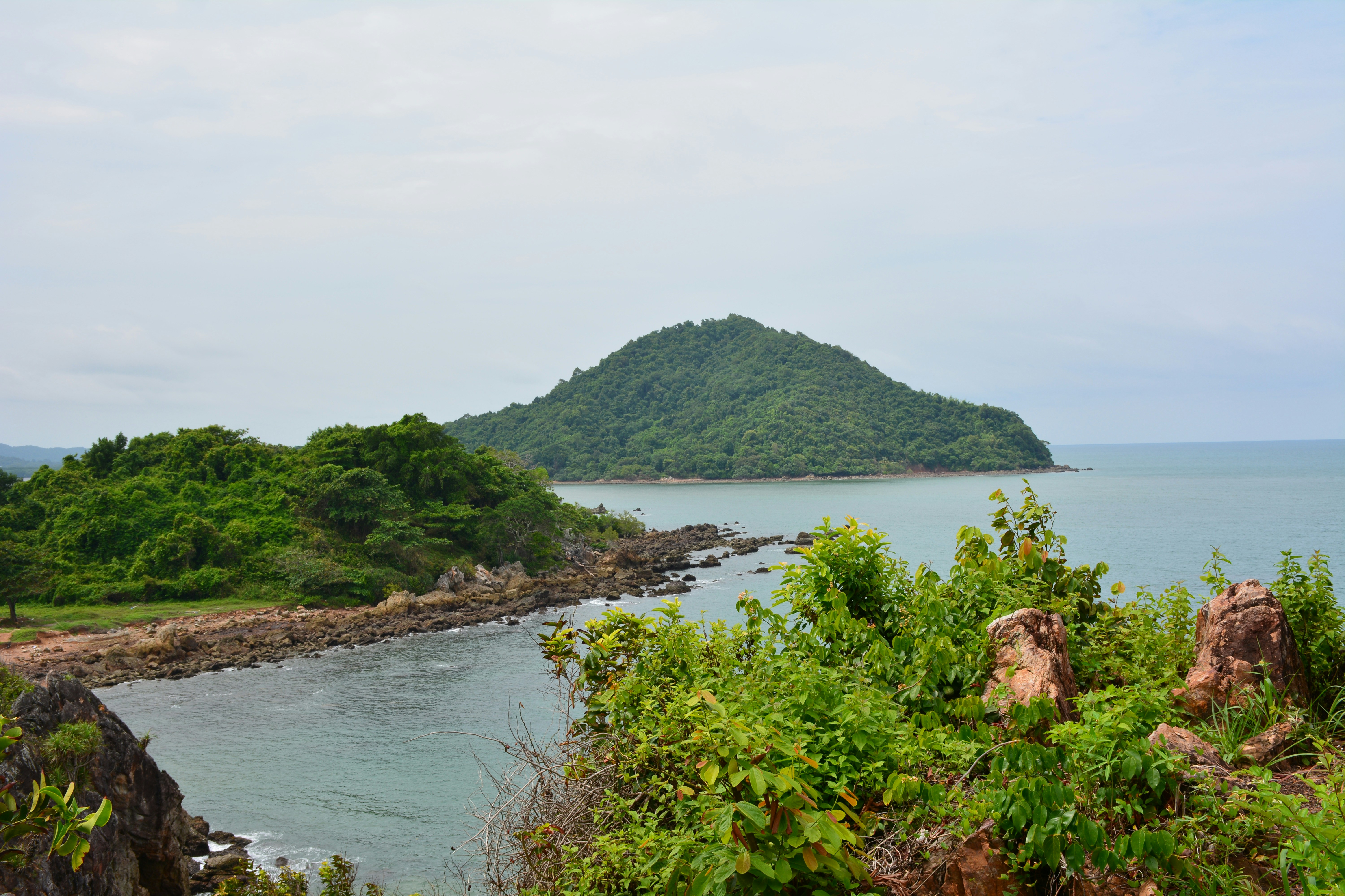 A lush island rises above calm coastal waters, framed by rocky foreground and green vegetation. A serene seascape emphasizing natural coastal scenery.