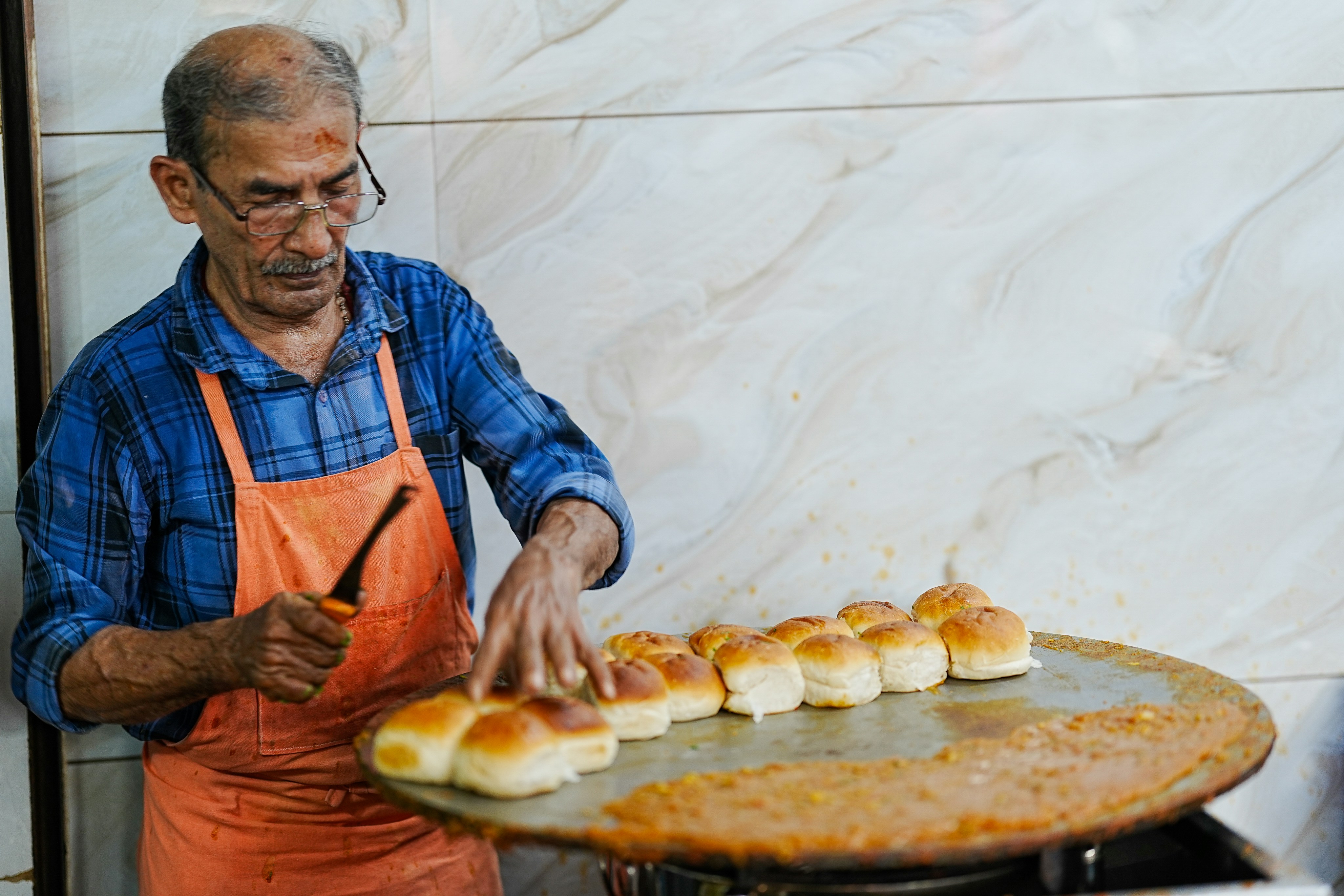 Baker in blue shirt and orange apron tends blistered buns on a large round griddle, slicing or shaping dough as heat radiates off the bread. A focused kitchen scene with warm tones.