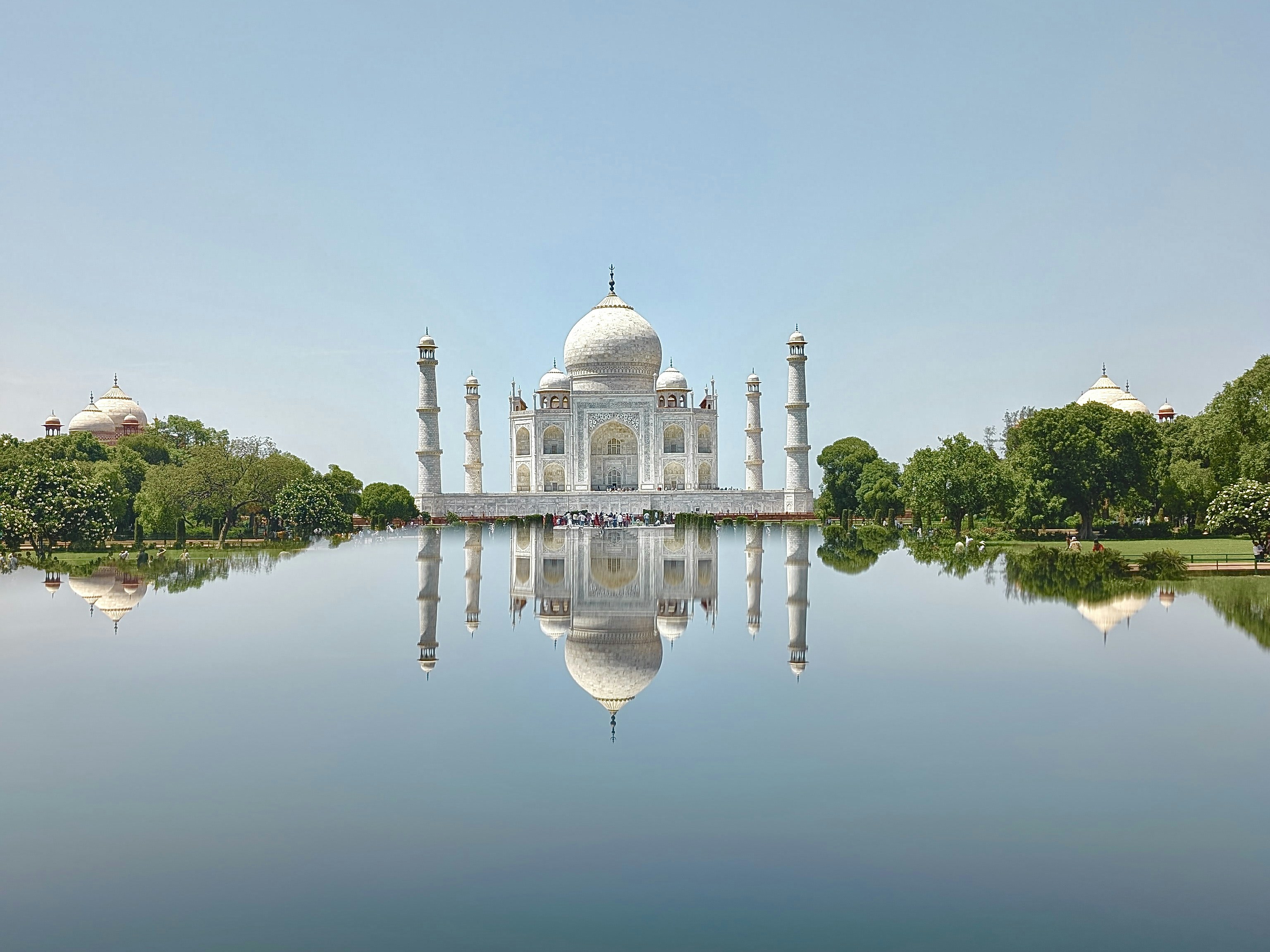 White marble mausoleum framed by reflecting pool with mirrored symmetry and surrounding gardens. Clear daytime lighting highlights the central dome and minarets with a calm, glassy water reflection.
