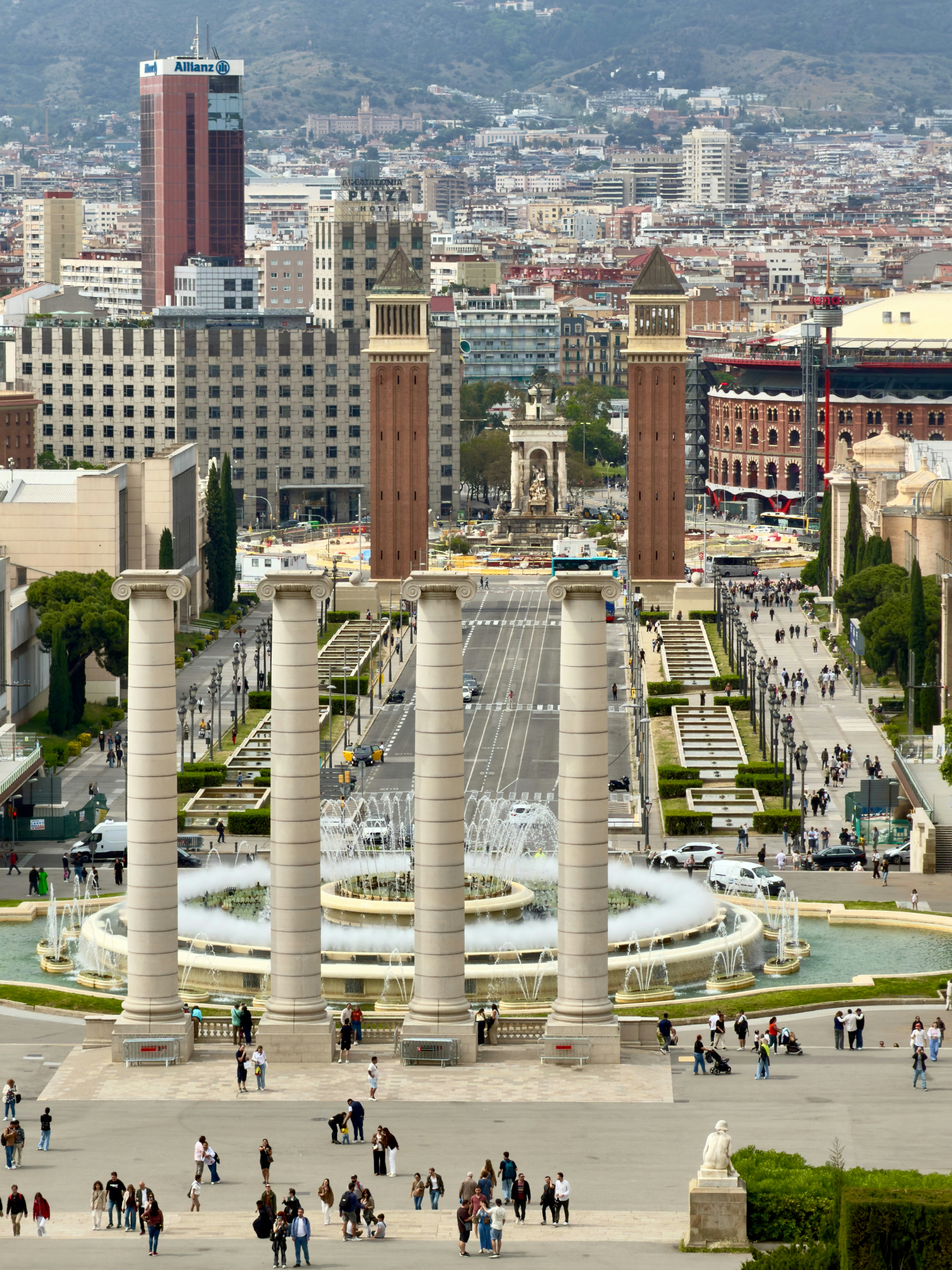 Wide view of the Montjuïc monumental plaza with a row of tall columns, cascading fountains, and a broad boulevard framed by historic and modern buildings. Pedestrians populate the foreground while Barcelona’s cityscape stretches beyond.