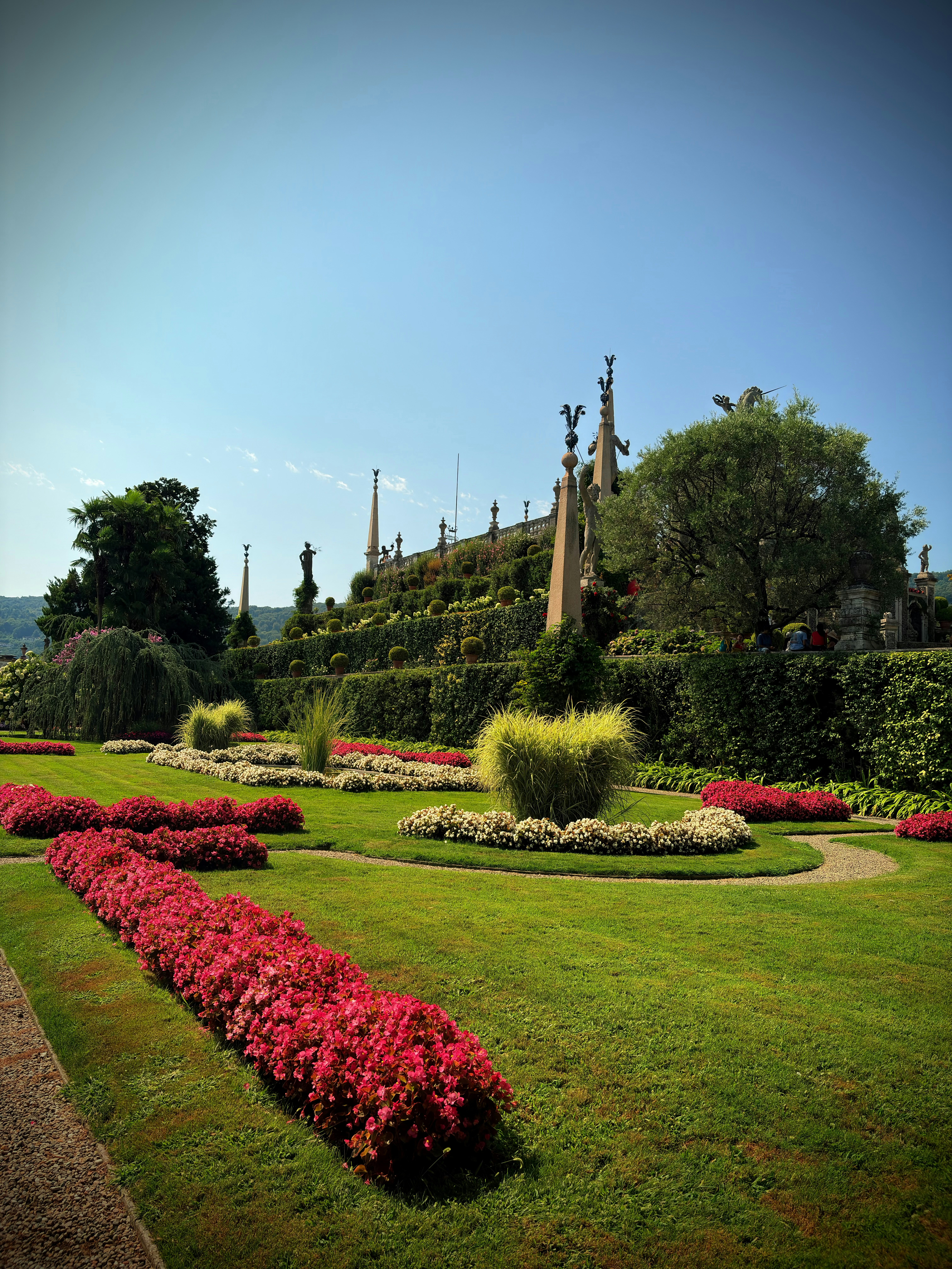 Colorful, manicured flowerbeds and geometric lawns frame a decorative terrace with statues and trimmed shrubs against a bright, open sky. A formal garden scene featuring vibrant pink and white blooms and lush greenery.