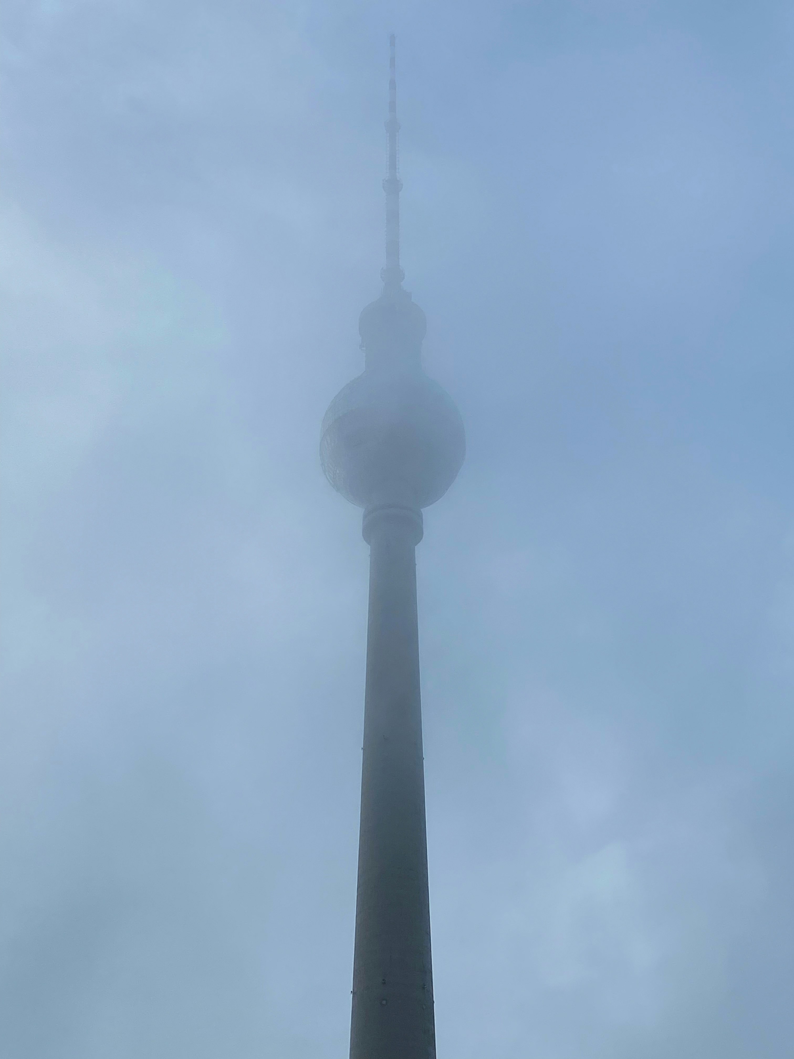 A tall communications tower rises into a hazy blue sky, softened by fog that blurs its silhouette. The scene emphasizes the vertical monument against a pale, atmospheric backdrop.