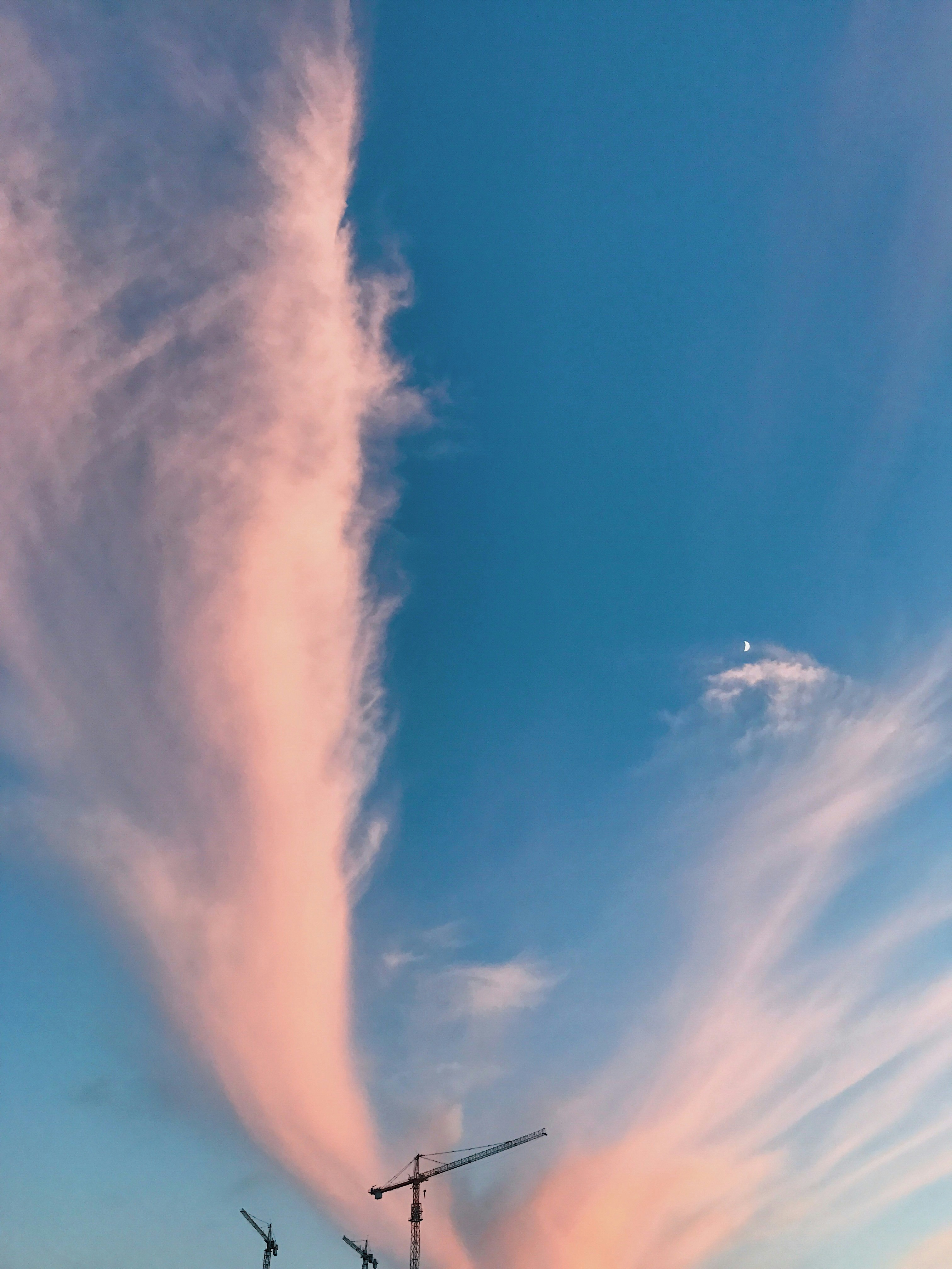 Soft pink-tinged clouds sweep across a blue sky above construction cranes, capturing a quiet, expansive early-evening atmosphere.