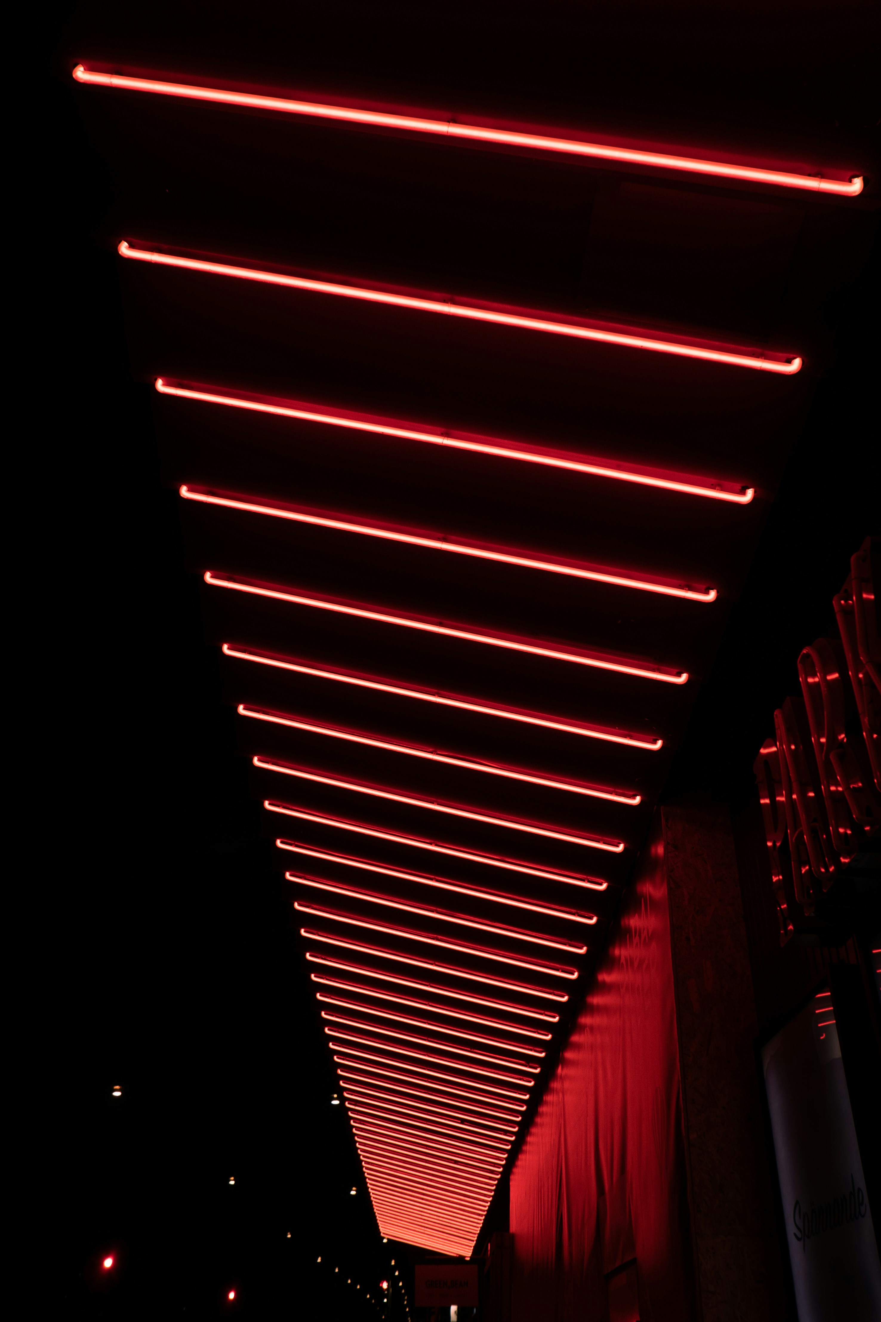 Red neon slats line a dim corridor, creating a dramatic, receding perspective with deep shadows and a moody atmosphere. A minimalist architectural scene with strong leading lines.
