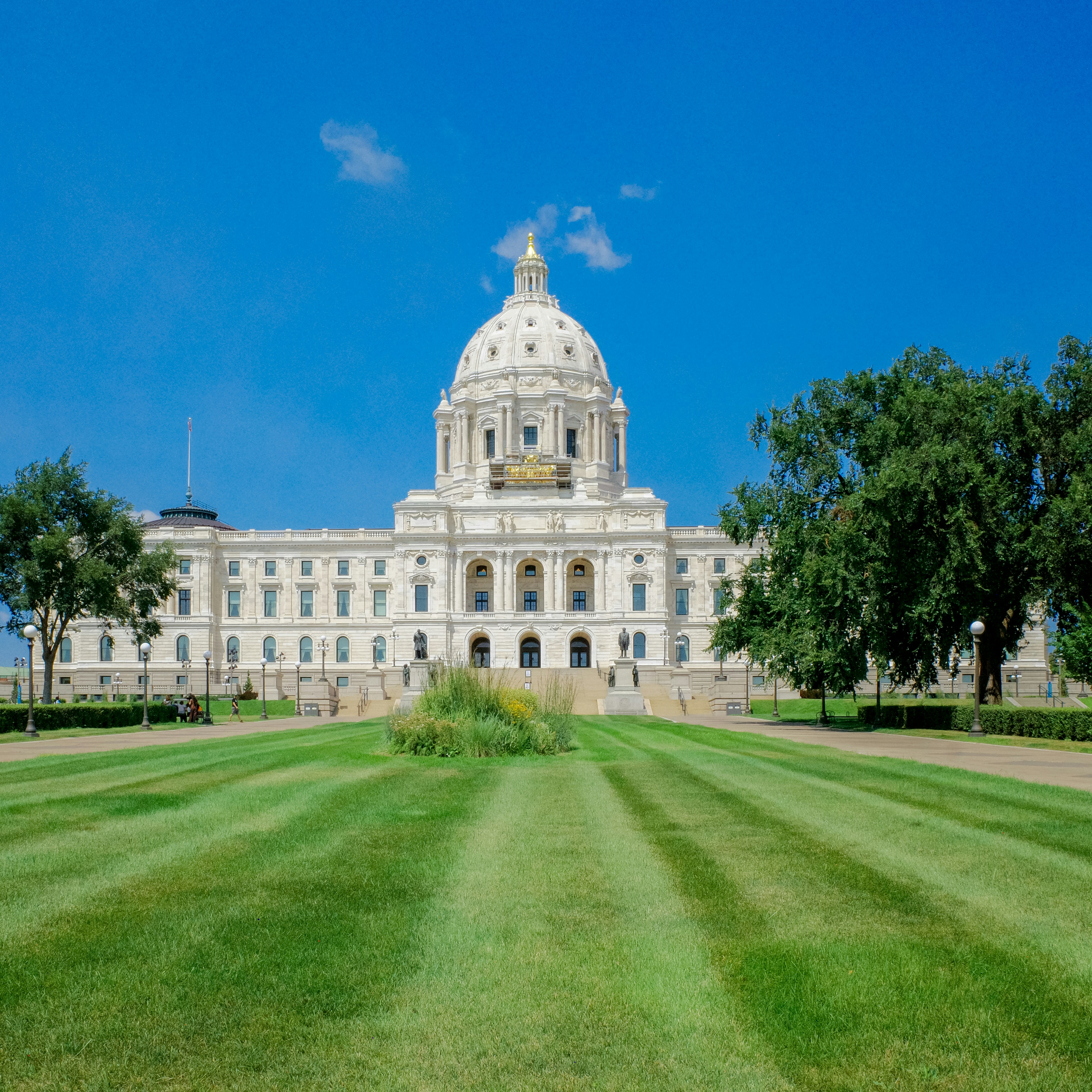 Sunlit capitol building rises behind a neatly striped green lawn; the scene emphasizes the architectural facade and expansive grounds.