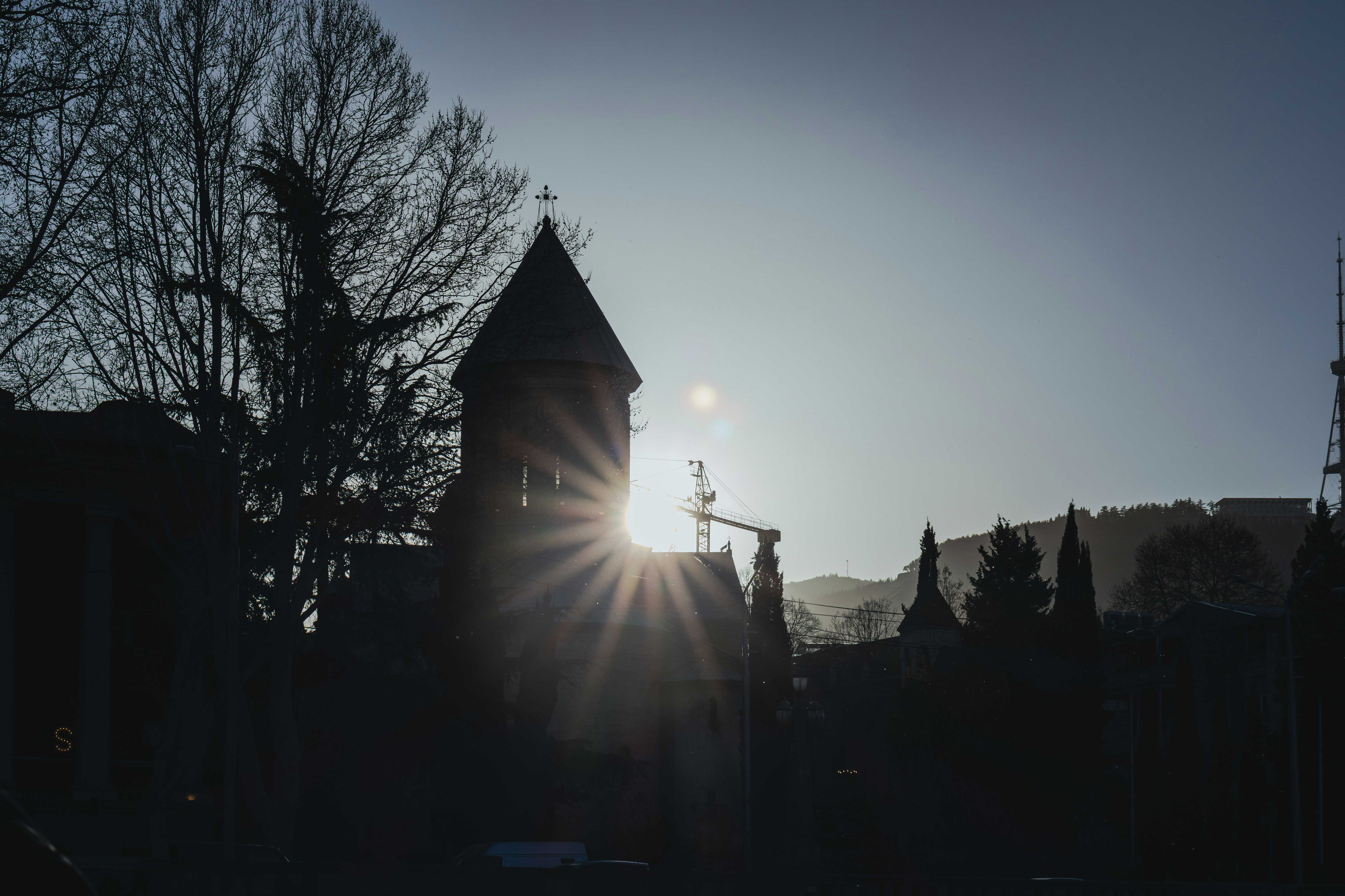 Silhouette of a cityscape at sunrise with a prominent tower center-left, sun rays radiating through the structure; a calm, low-contrast scene emphasizing architectural forms.