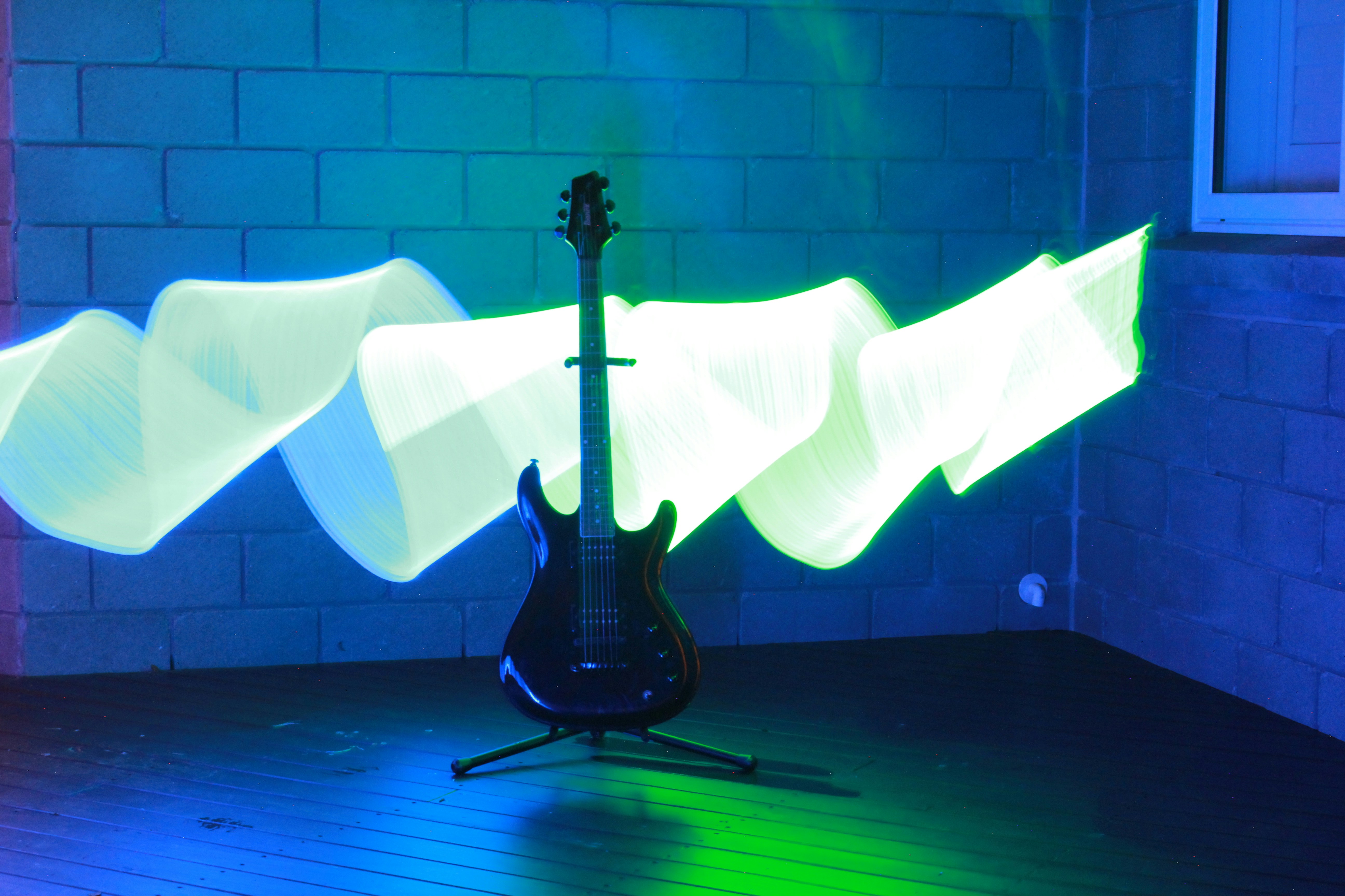 Electric guitar on a stand in a dim room, illuminated by blue and green light trails swirling behind it, with a textured brick wall backdrop.