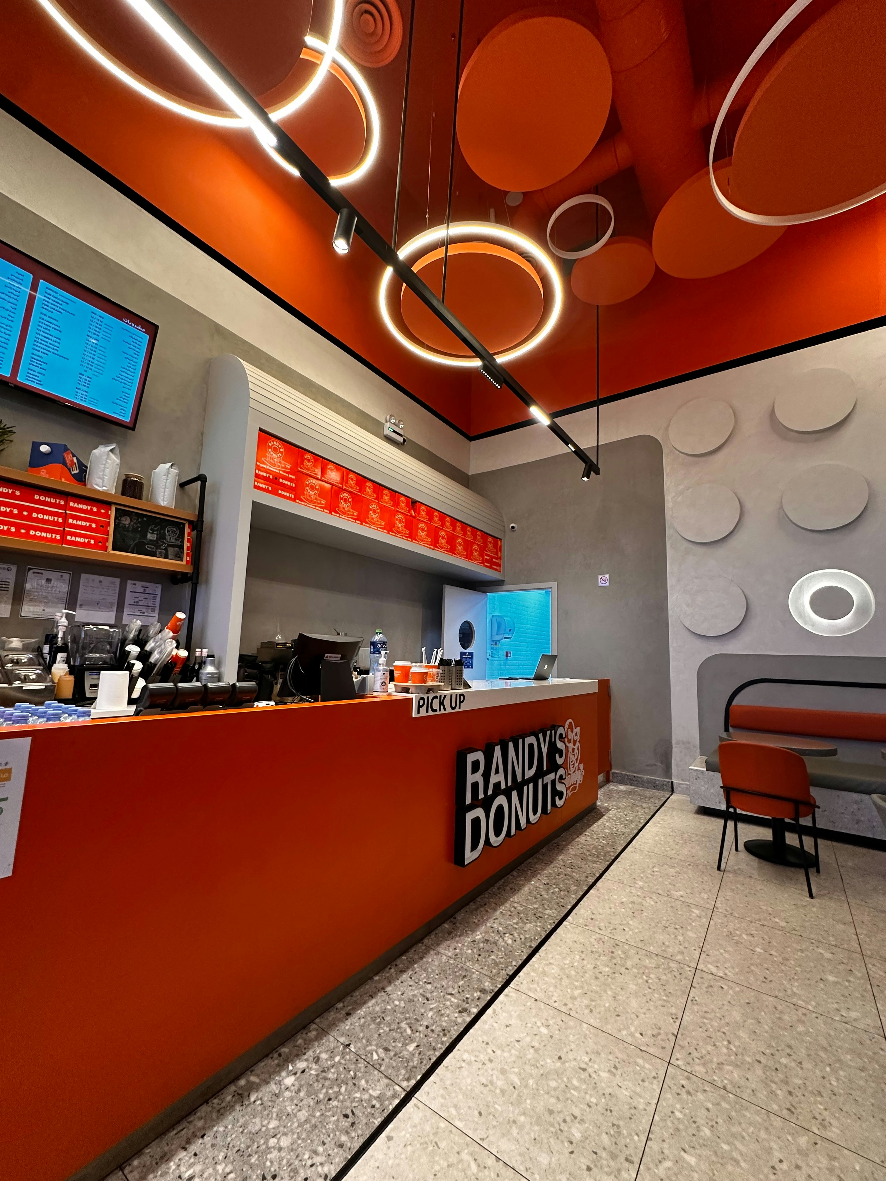 Sleek fast-casual counter interior with orange accents and circular ceiling lamps; front counter reads 'RANDY'S DONUTS' and a pickup area is visible in a modern dining space.