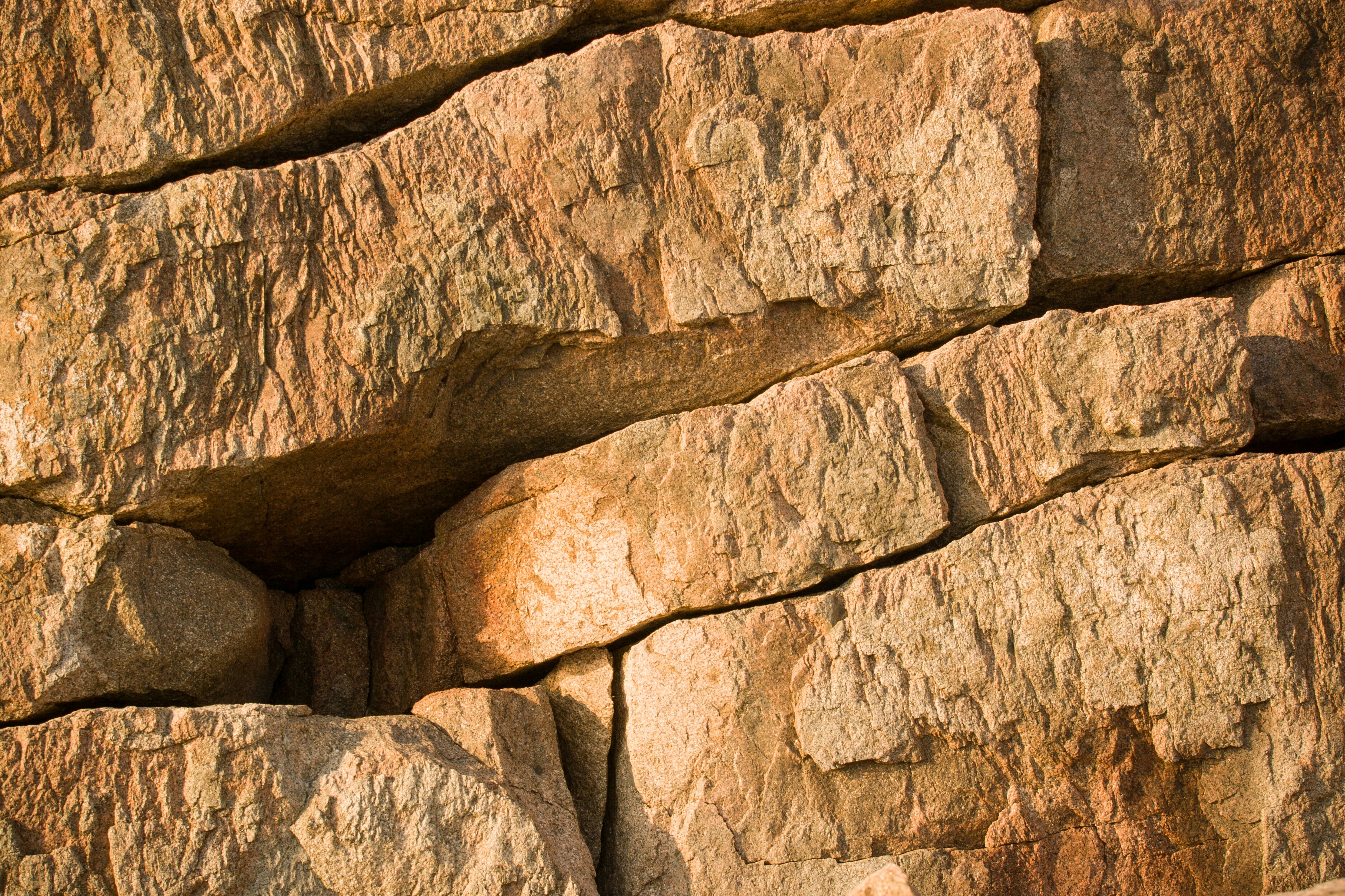 Sunlit textured stone wall with layered granite blocks showing warm, earthy tones and pronounced edge detail. A close-up of masonry texture highlighting natural roughness and subtle color variation.