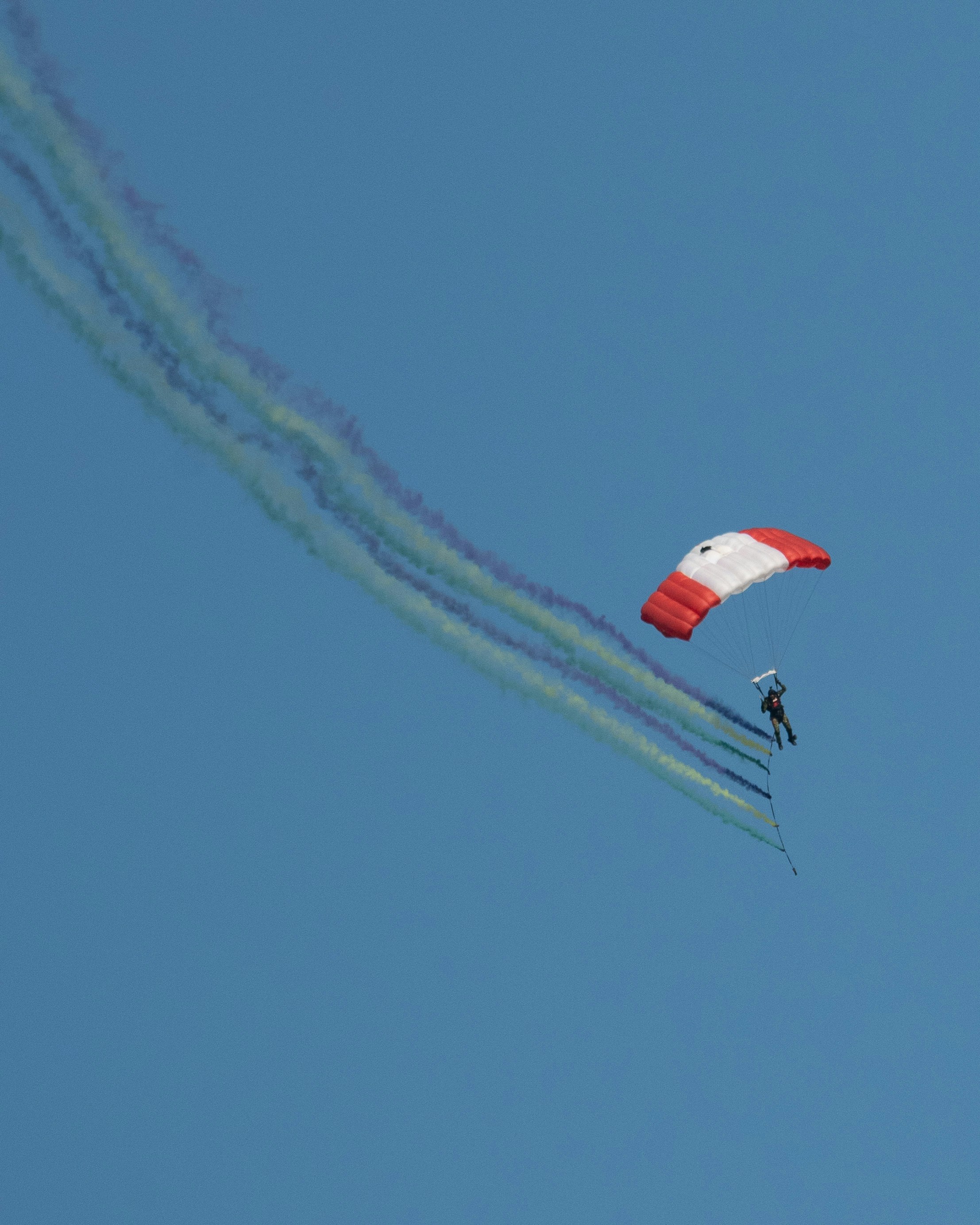 Parachutist trailing colored smoke arcs across a clear blue sky, parachute open and lines streaming behind, isolated against the vast backdrop. Logo-like markings are minimal/absent; overall scene captures motion and technique.
