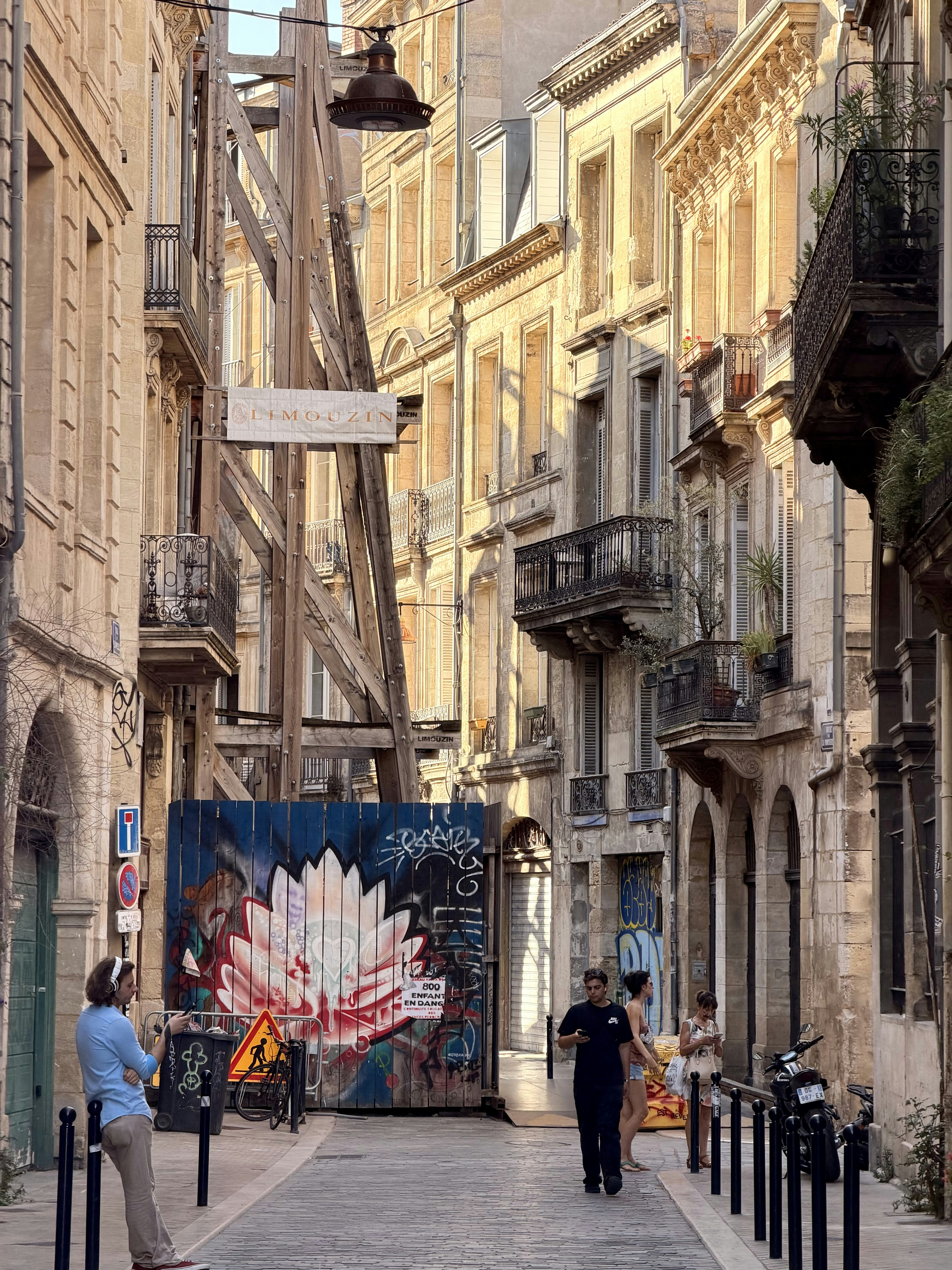 Sunlit alleyway with graffiti gate and people strolling under warm stone façades
