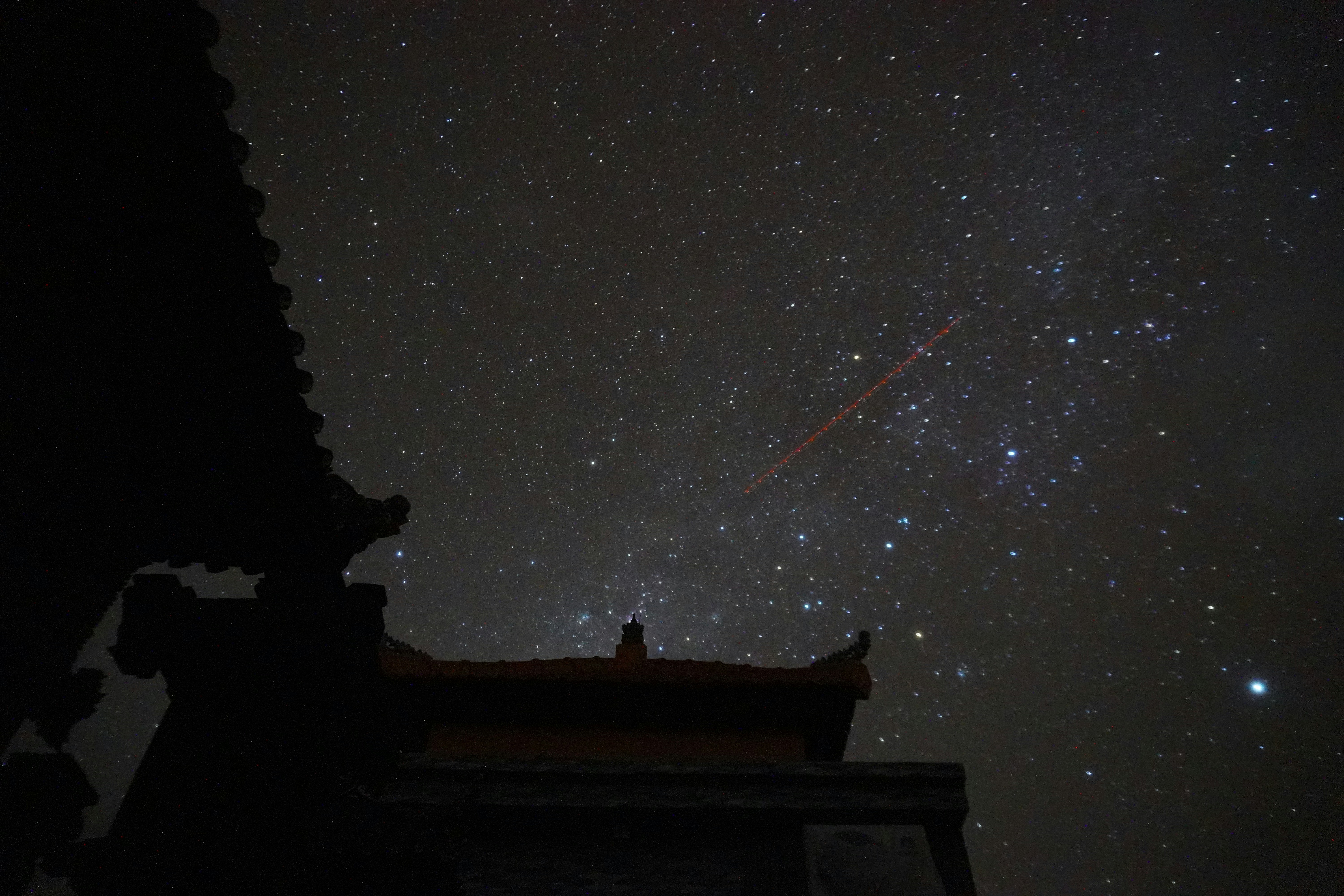 Night sky filled with stars above a silhouetted temple roof; a faint meteor streaking the stars adds motion to the tranquil scene.