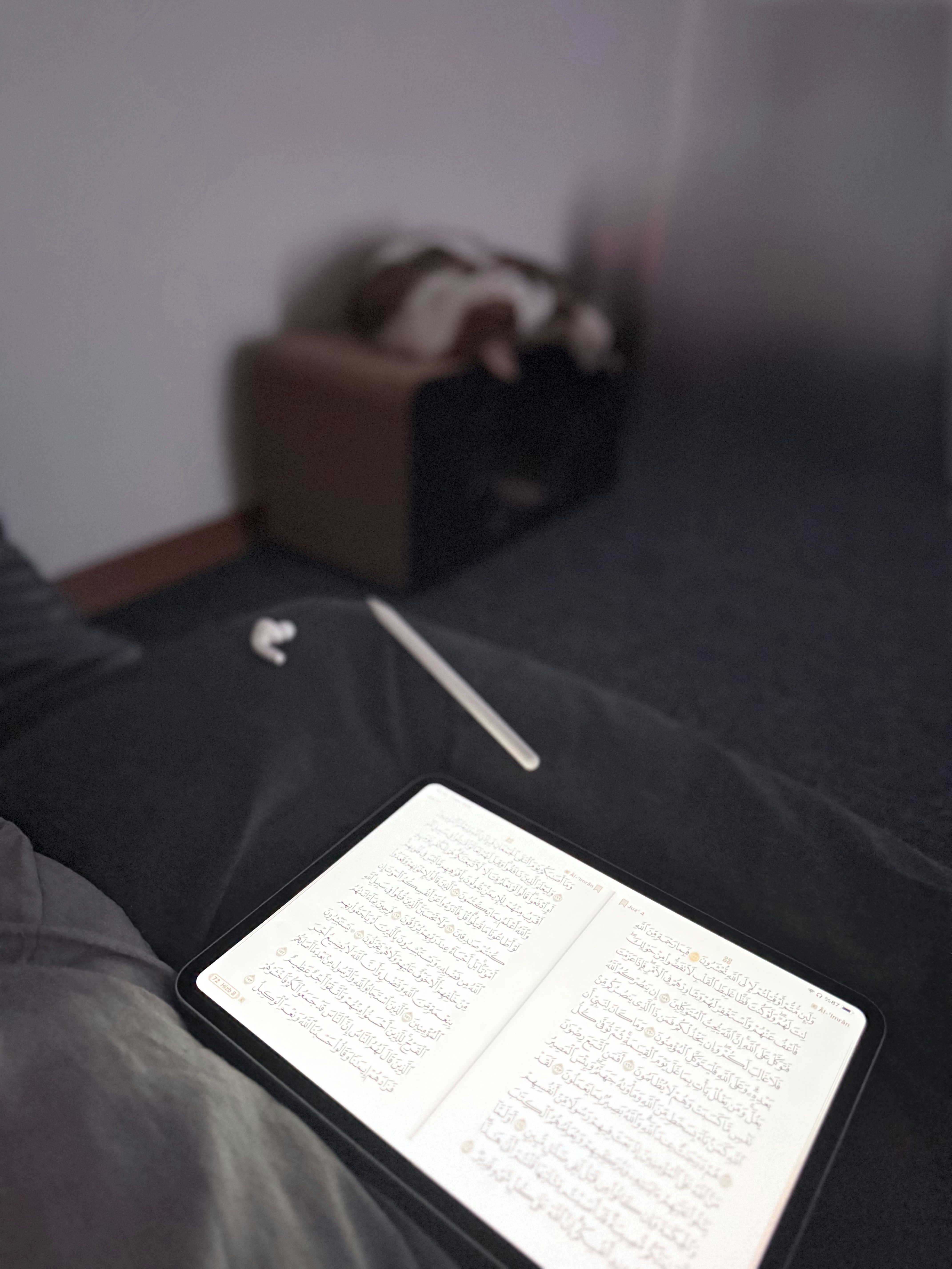 Arabic text displayed on an open tablet rests on a dark bed, with a stylus nearby and a blurred living room in the background.