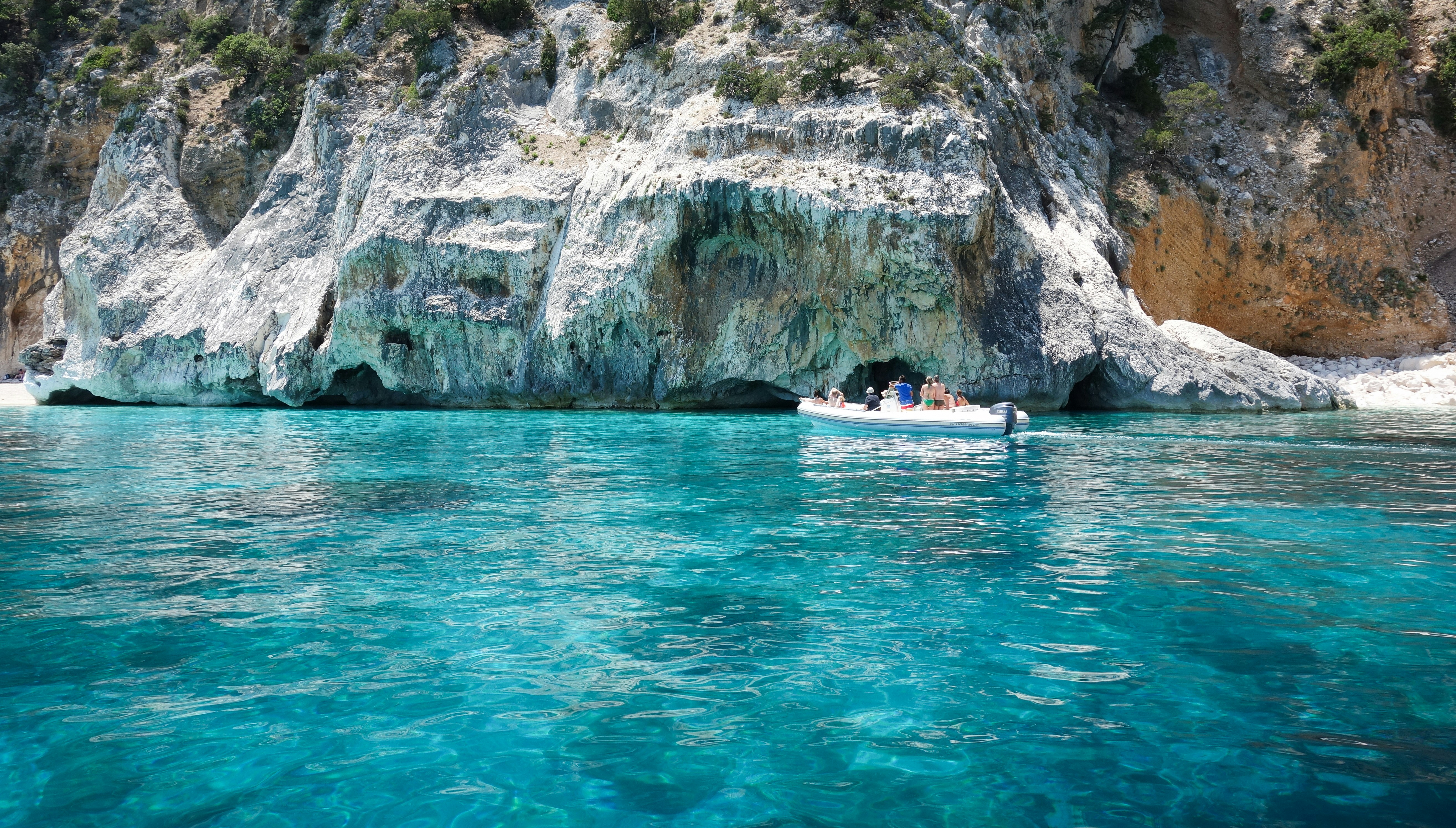 Bright turquoise sea meets a rugged limestone cliff with a small boat near a dark cave opening.