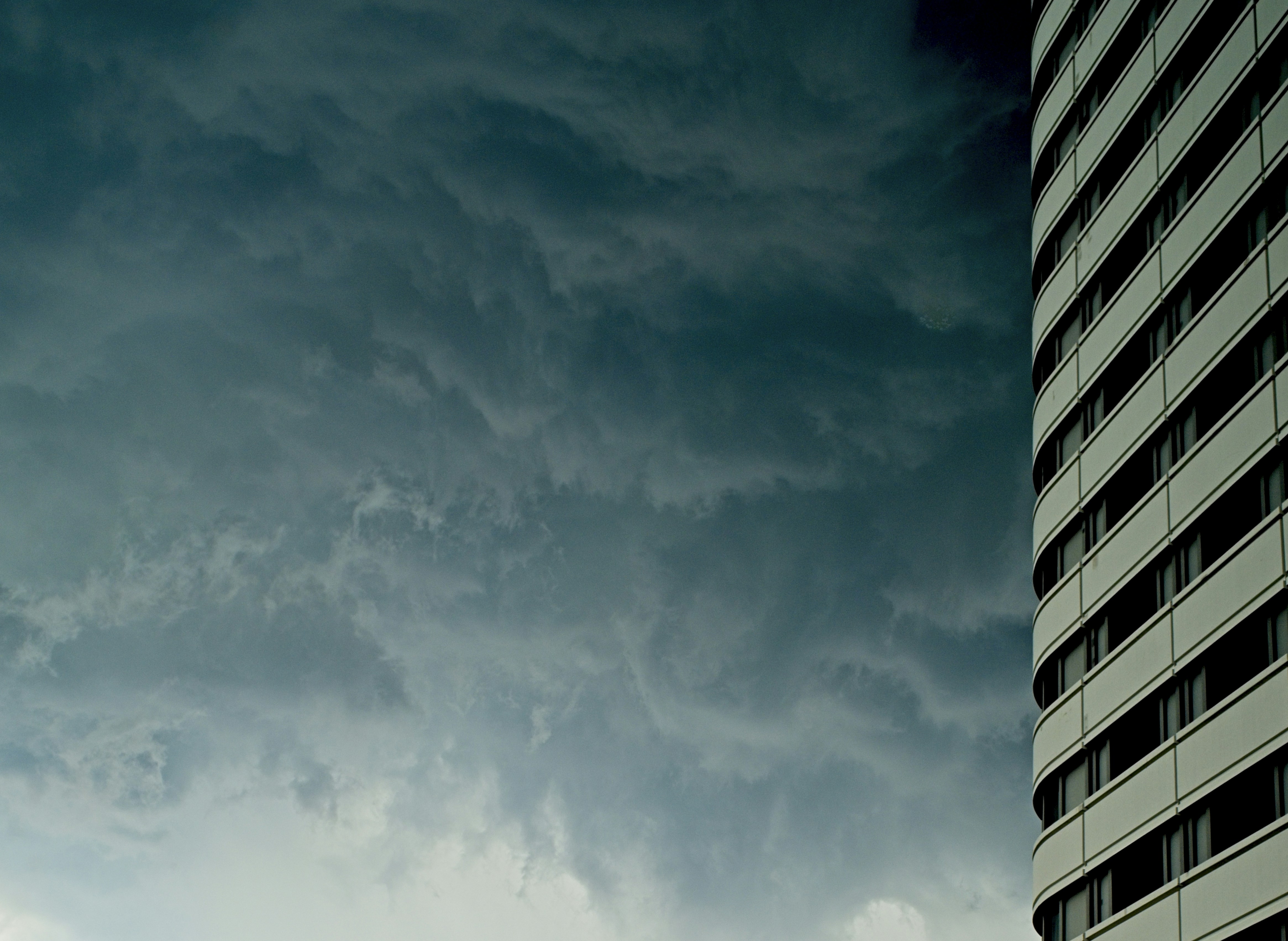Heavy, textured storm clouds dominate the frame in deep shades of gray and blue, their rolling forms swirling ominously above a solitary curved hotel tower at the frame's edge. The building's repeating rows of windows add a stark architectural contrast to the moody, atmospheric sky. A tiny distant bird or object punctuates the vast expanse of cloud above. The minimalist composition captures the brooding calm just after a summer thunderstorm has passed through the city.