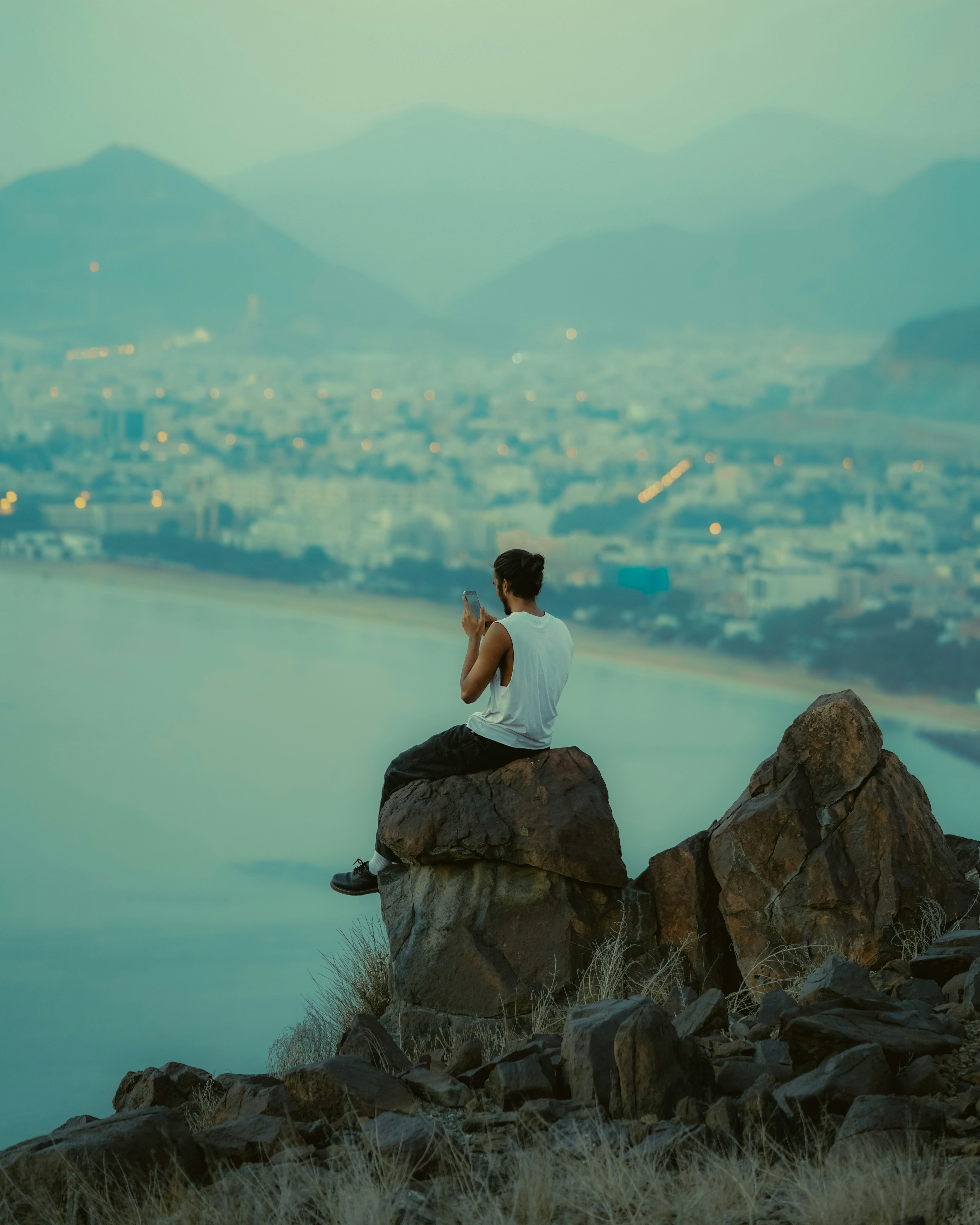 A lone photographer sits on rugged rocks overlooking a coastal city as mountains loom in the distance; the scene emphasizes solitude and perspective near dusk.