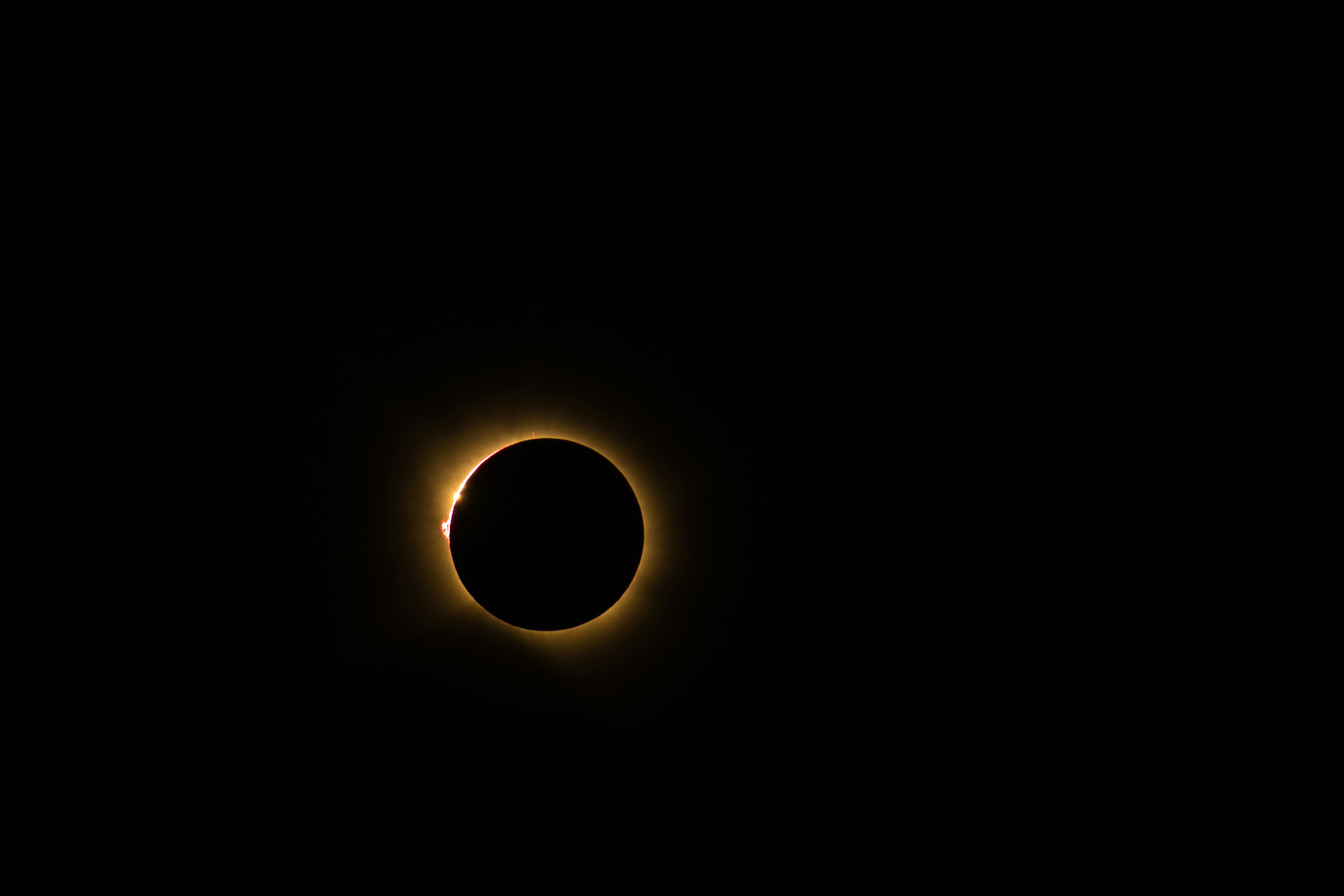 A near-dark solar eclipse reveals a glowing corona around a black disk, with a hint of solar prominence on the edge, isolated against a black sky.