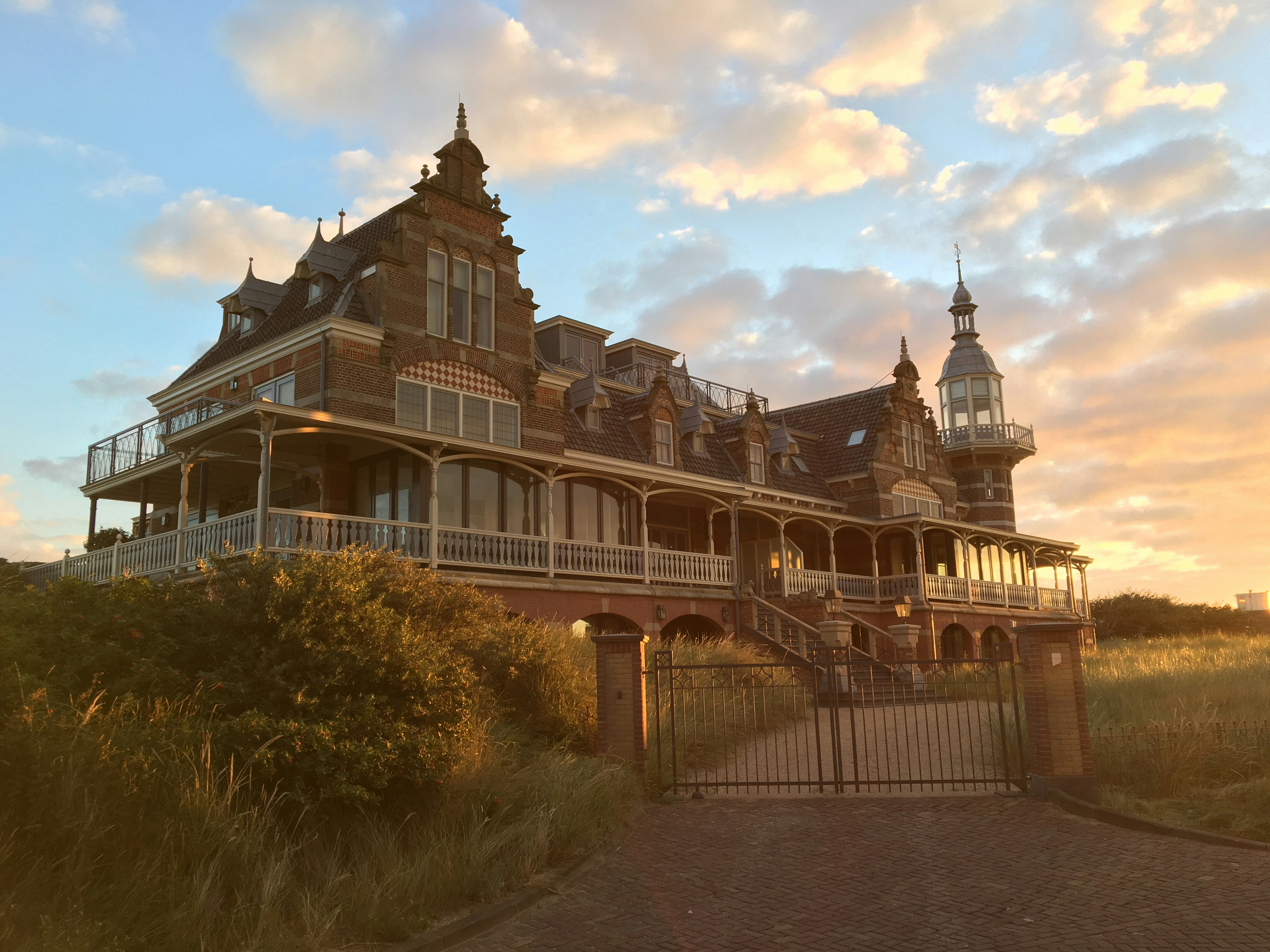 Elegant cliffside mansion with expansive wraparound veranda bathed in warm sunset light; foreground iron gate and tall grasses lead to the grand wooden structure.