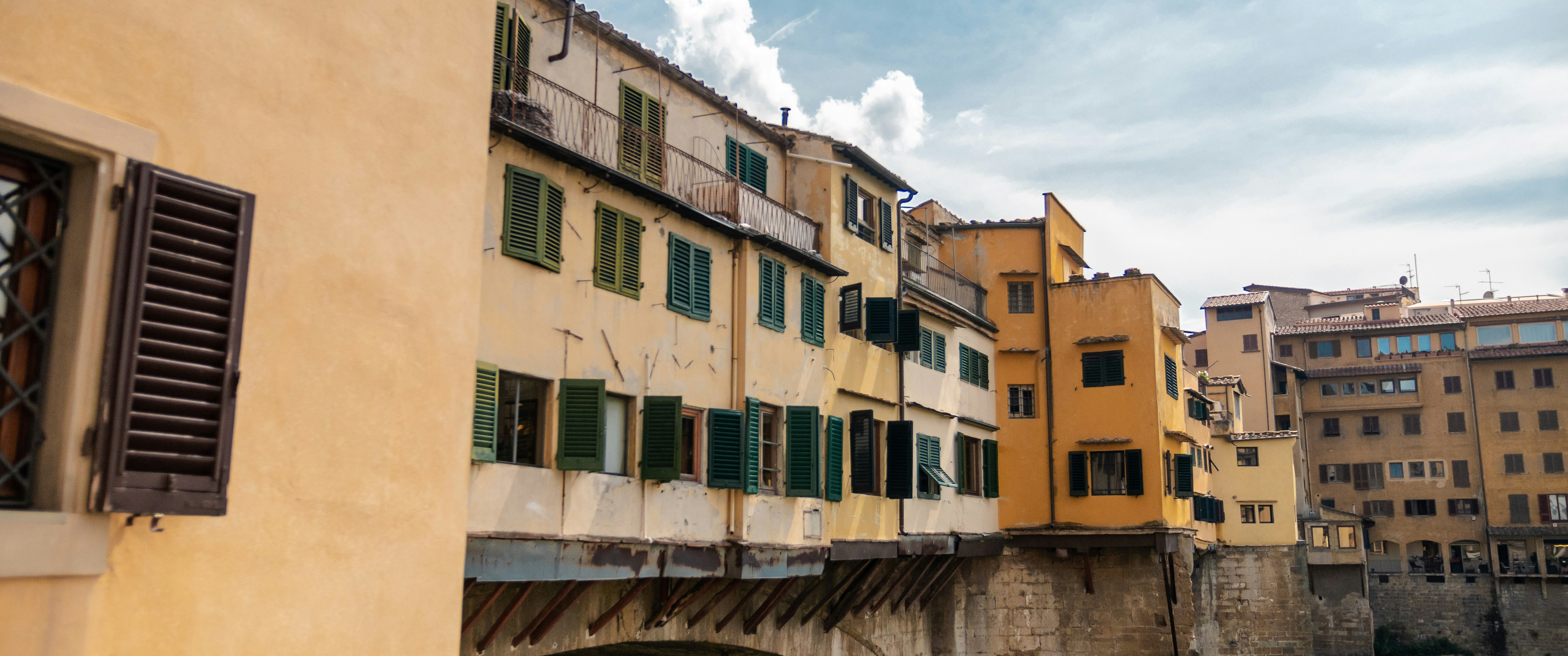 Colorful town façades line a sunlit riverbank with shutters open and reflections on the water, conveying a quaint, coastal European streetscape.