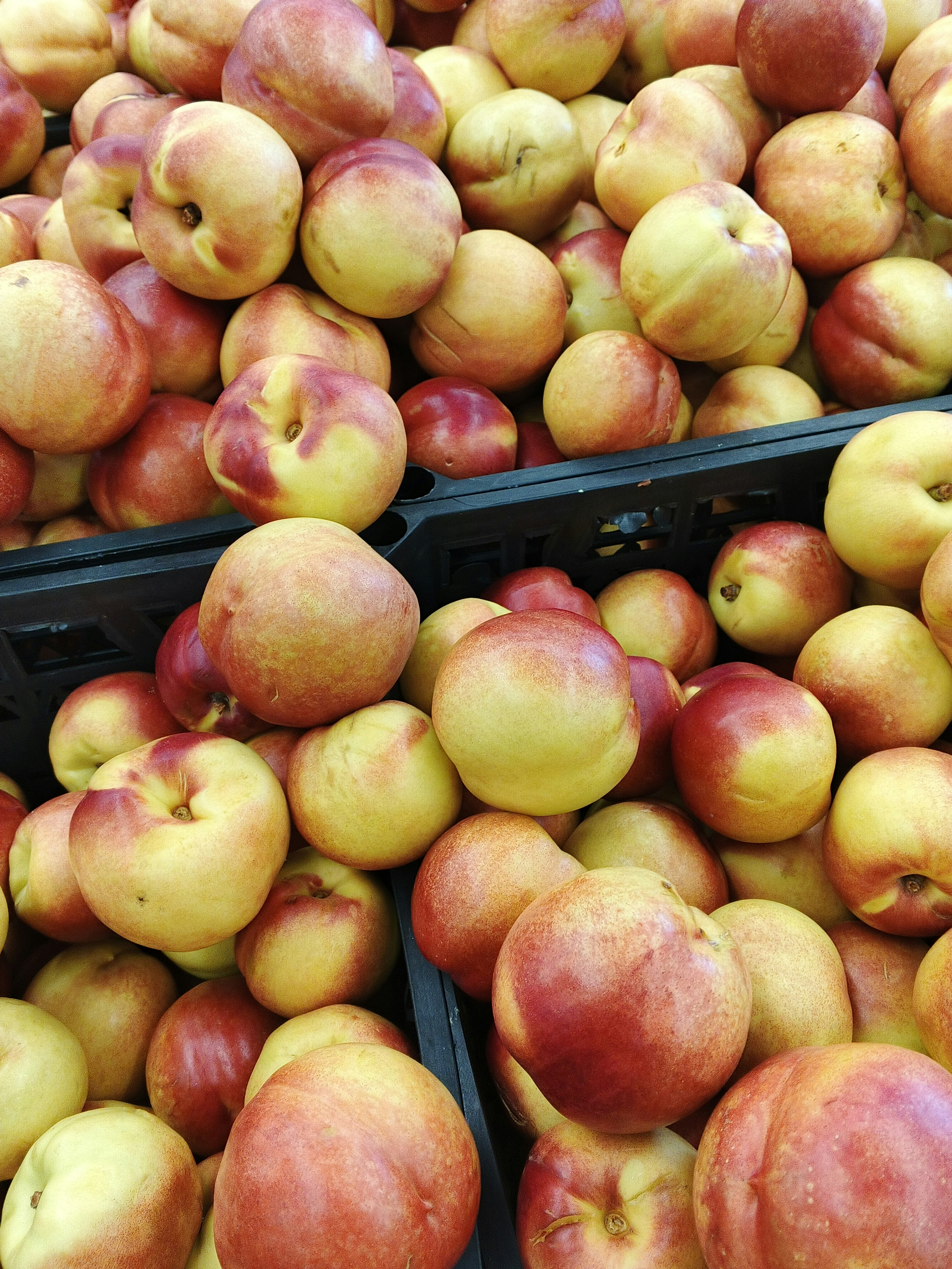 Pile of yellow-red peaches packed in black crates, close-up emphasis on fruit textures and color variety. Logo/branding not present.