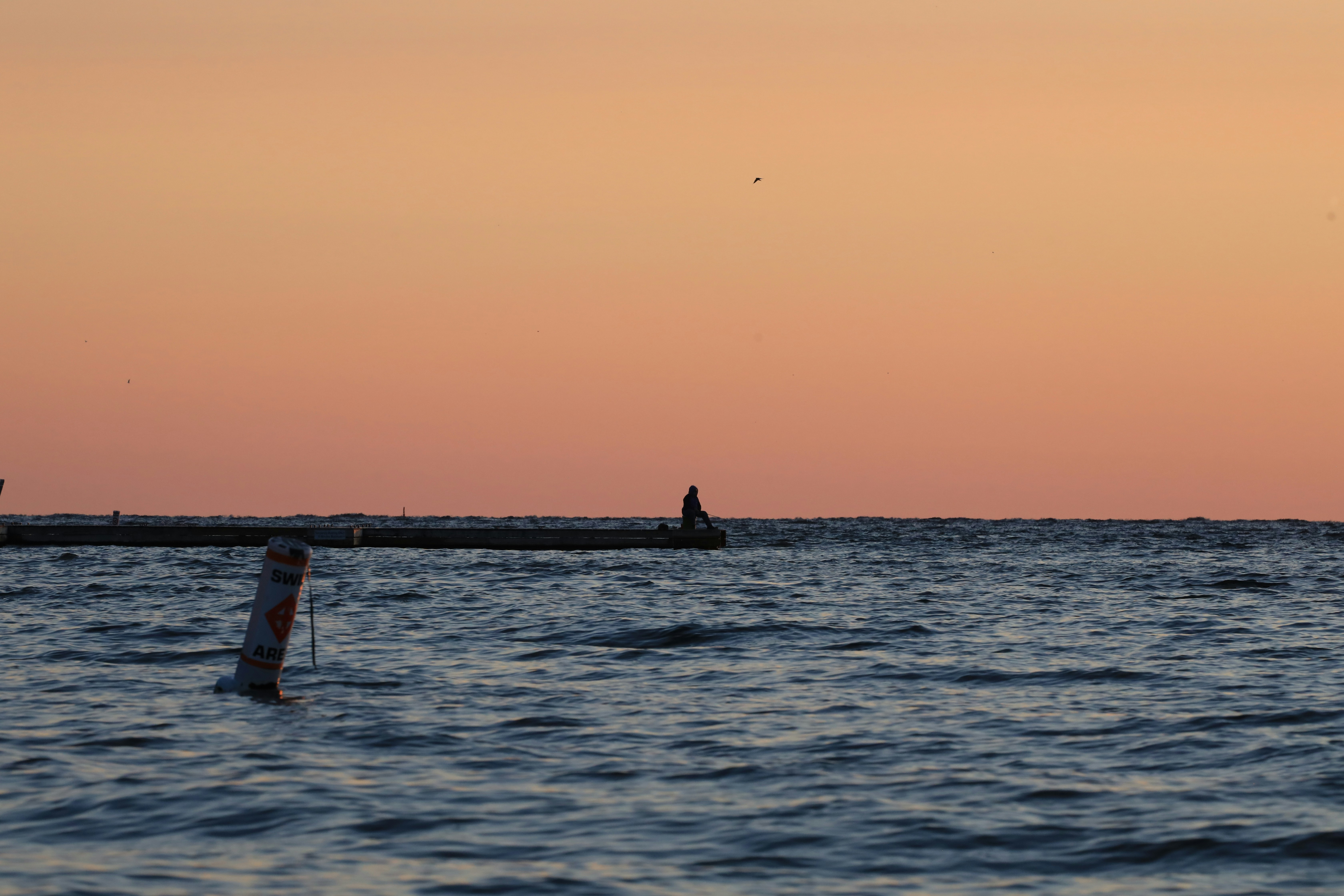 Silhouetted figure on a jetty at sunset over calm waves, with a buoy in the foreground and a warm colored sky. A tranquil coastal scene; a photograph capturing quiet moments at sea.