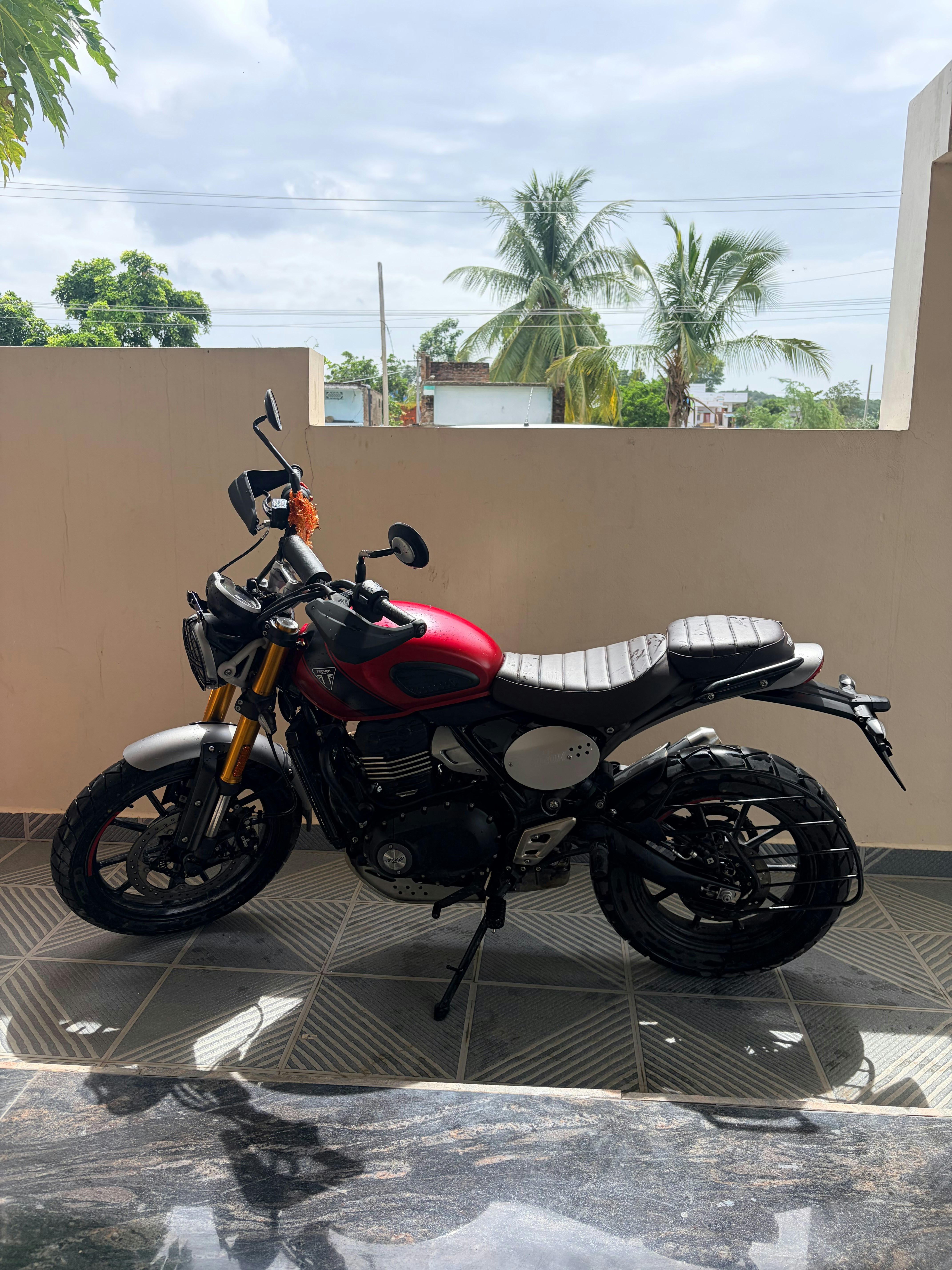 Red motorcycle parked in a sunlit courtyard against a beige wall, with palm trees visible over the top. The image captures the bike’s chrome and matte surfaces with strong mid-day light.
