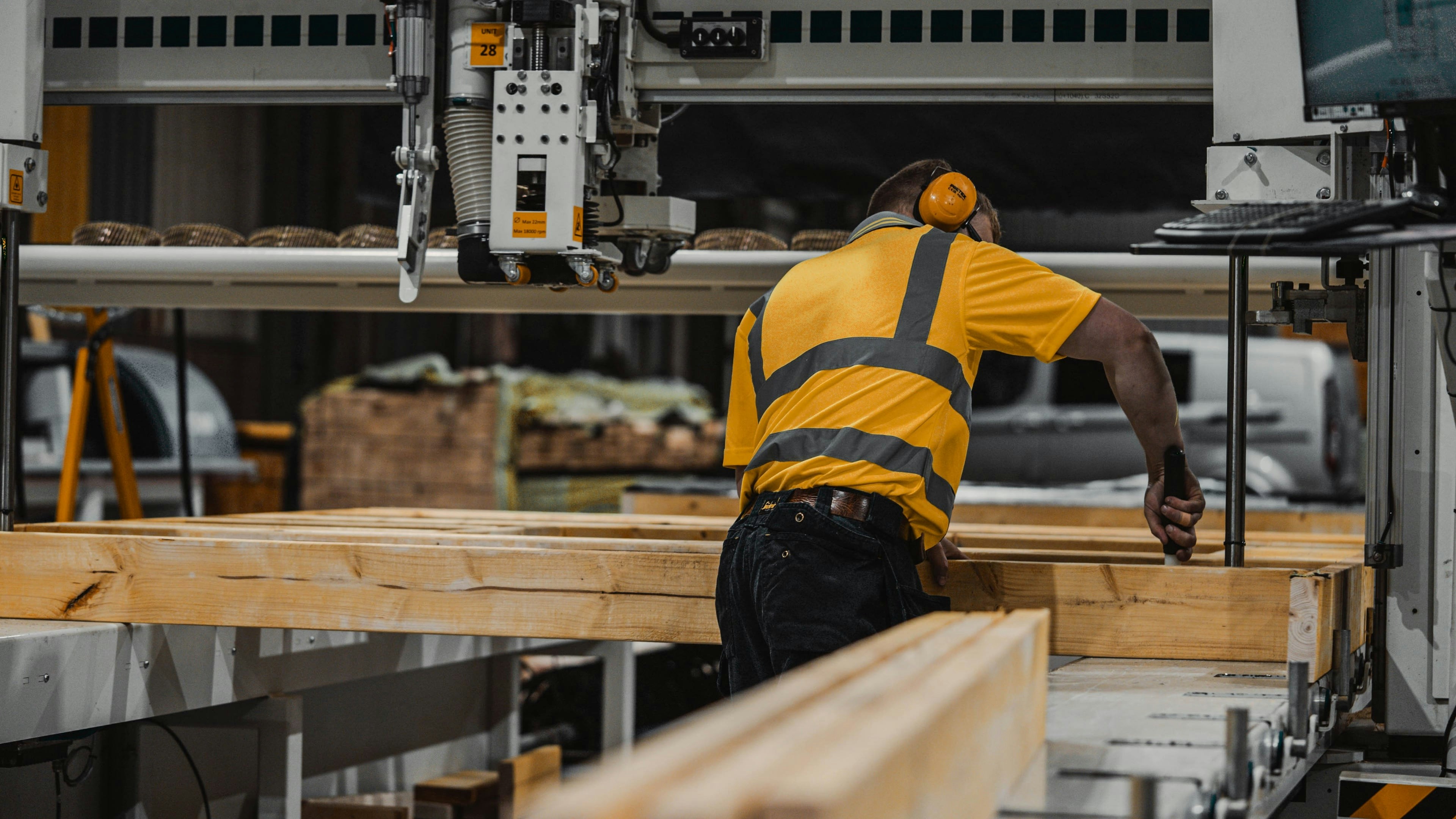 Worker in a high-visibility vest guides a wooden beam beneath industrial machinery in a wood processing facility.