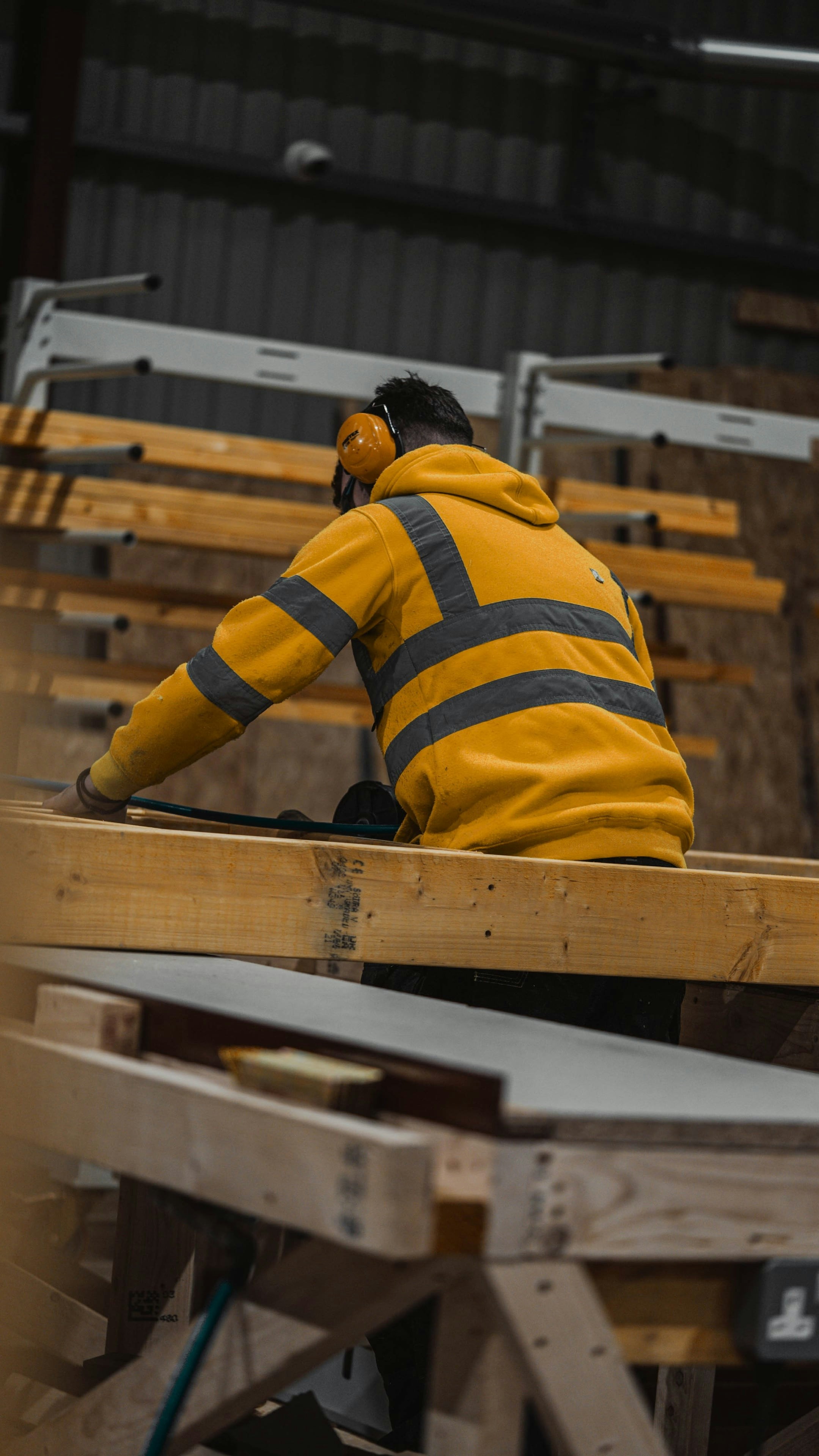 Construct worker in a bright yellow safety jacket and earmuffs handling timber at a workshop; a close-up of stacked planks and carpentry work inside a dimly lit industrial space.