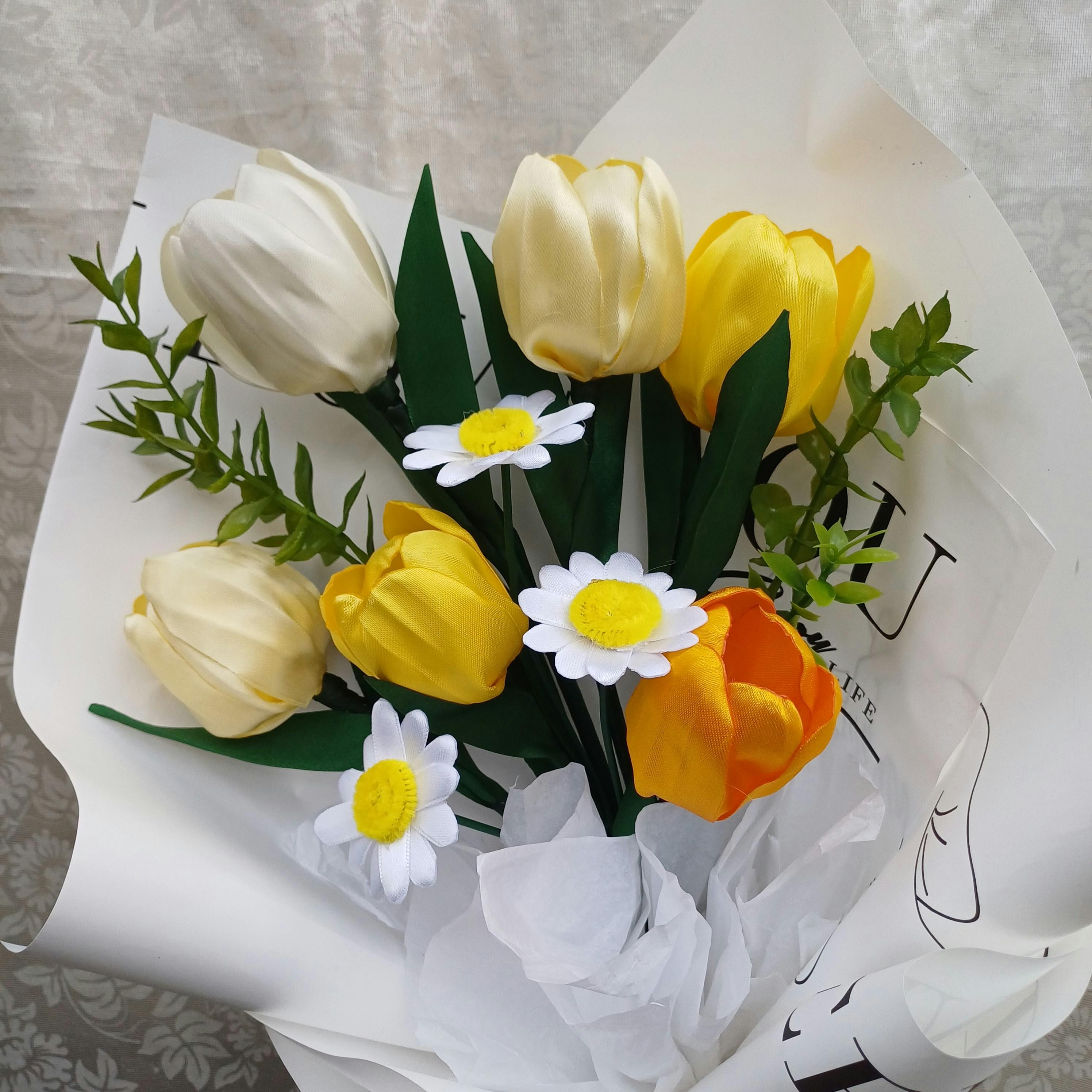 Bouquet of cream and yellow tulips with white daisies, wrapped in white paper. A floral arrangement shot with soft lighting and subtle texture in the background.