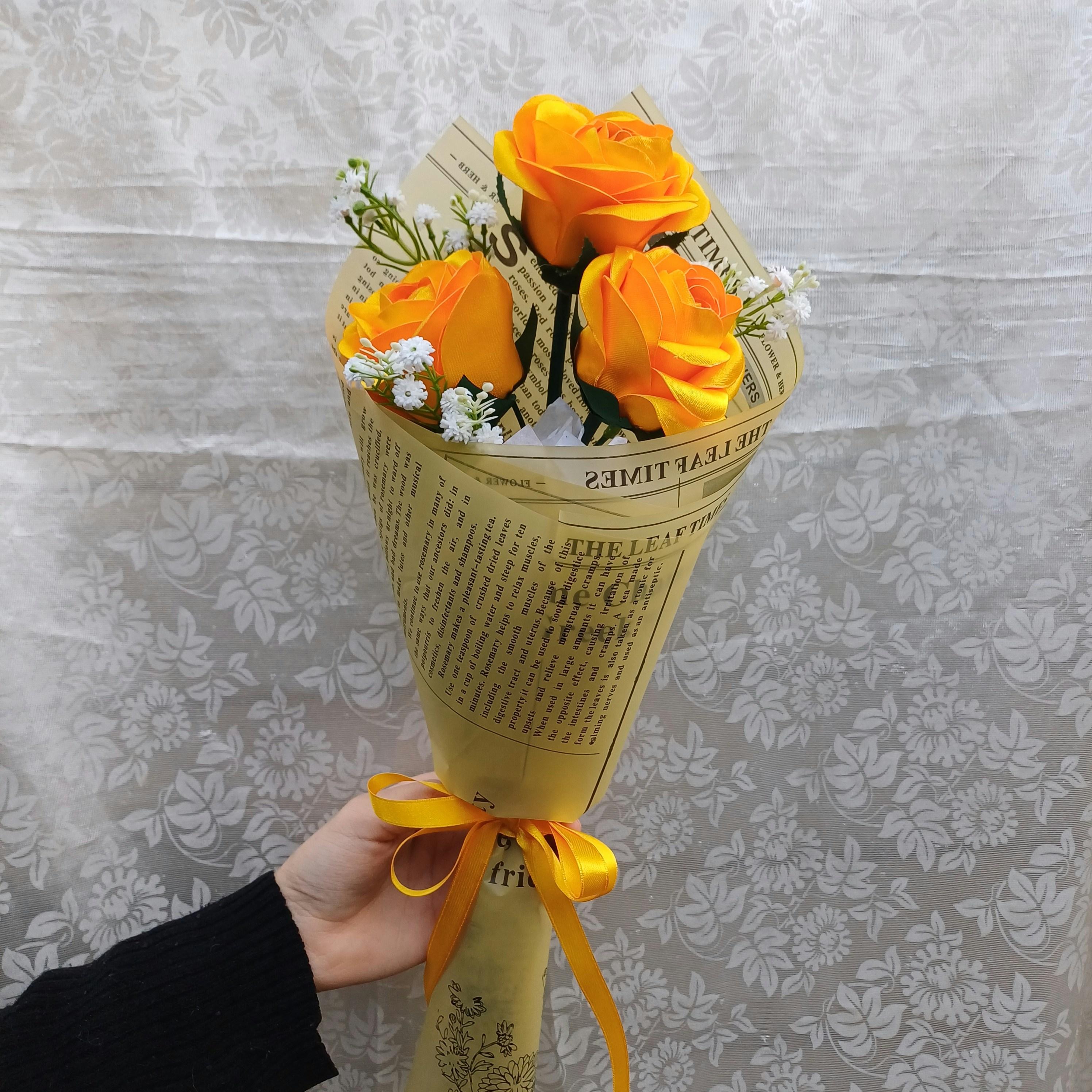 Bright orange roses wrapped in newspaper-style paper with a yellow ribbon, held by a gloved hand against a patterned backdrop.