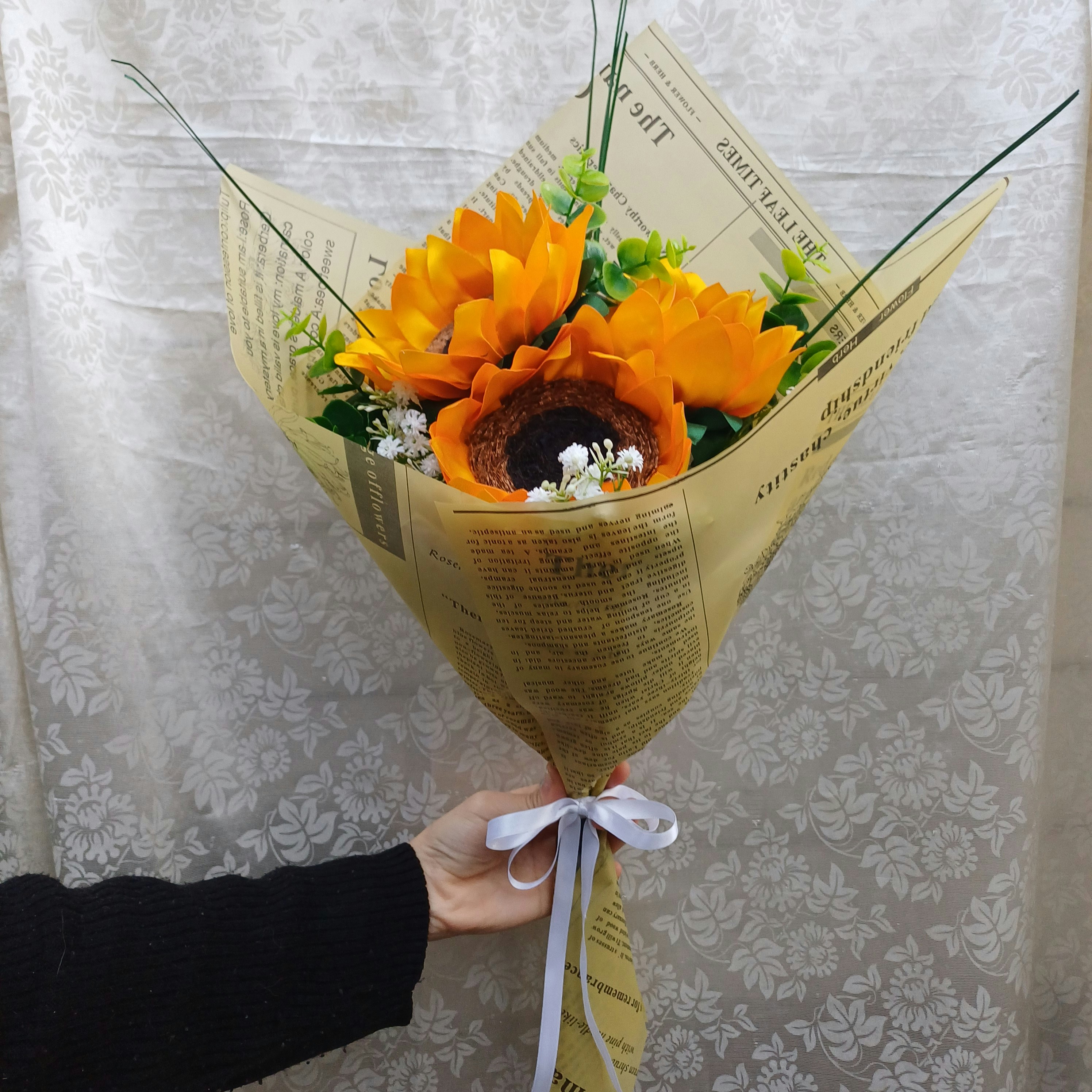 Bright bouquet of orange sunflowers wrapped in newspaper, held by a gloved hand against a pale patterned backdrop.