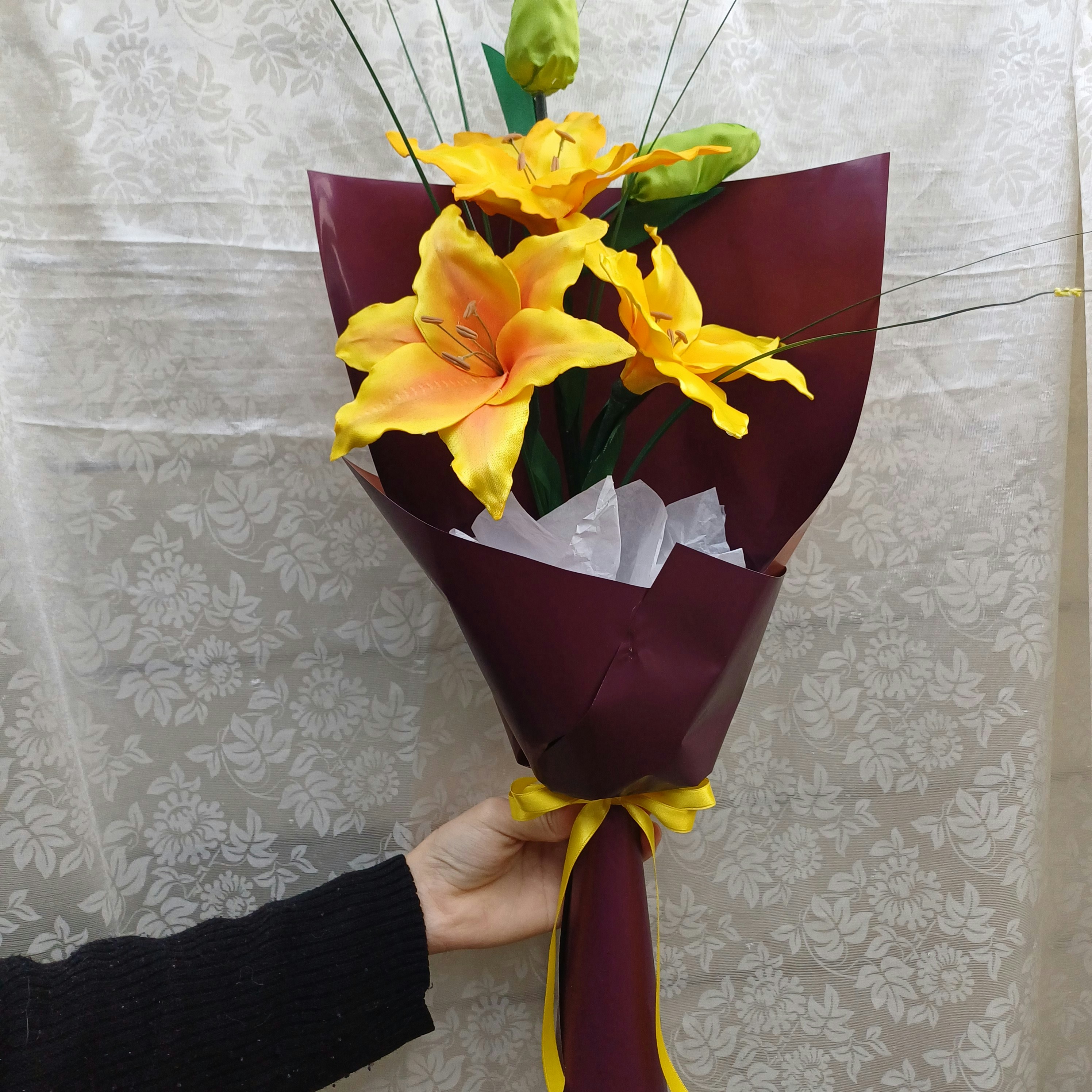 Vibrant yellow lilies arranged in a burgundy bouquet, held by a gloved hand against a patterned backdrop.
